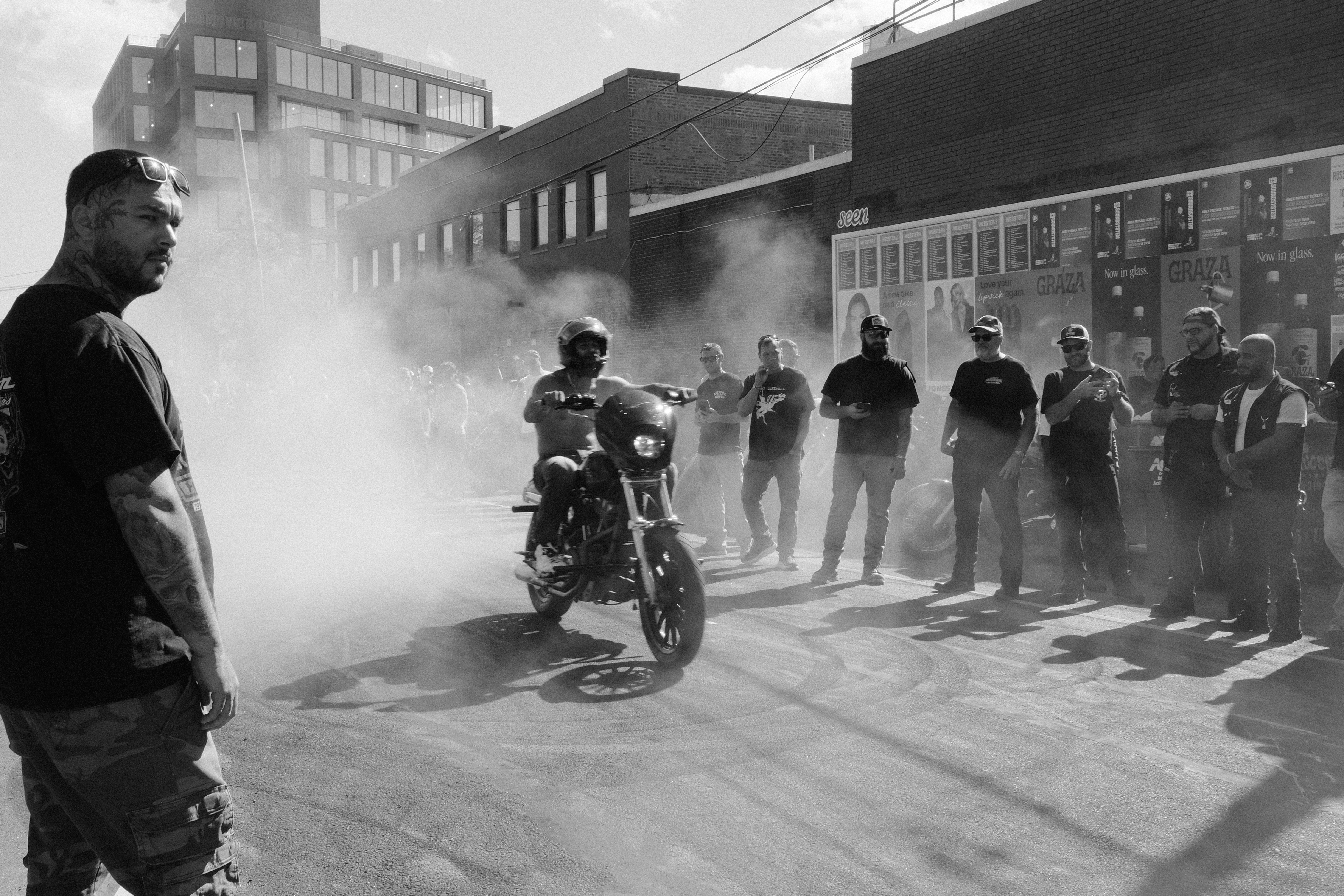 Biker performing a burnout amidst a crowd of onlookers, enveloped in a cloud of smoke on a city street.