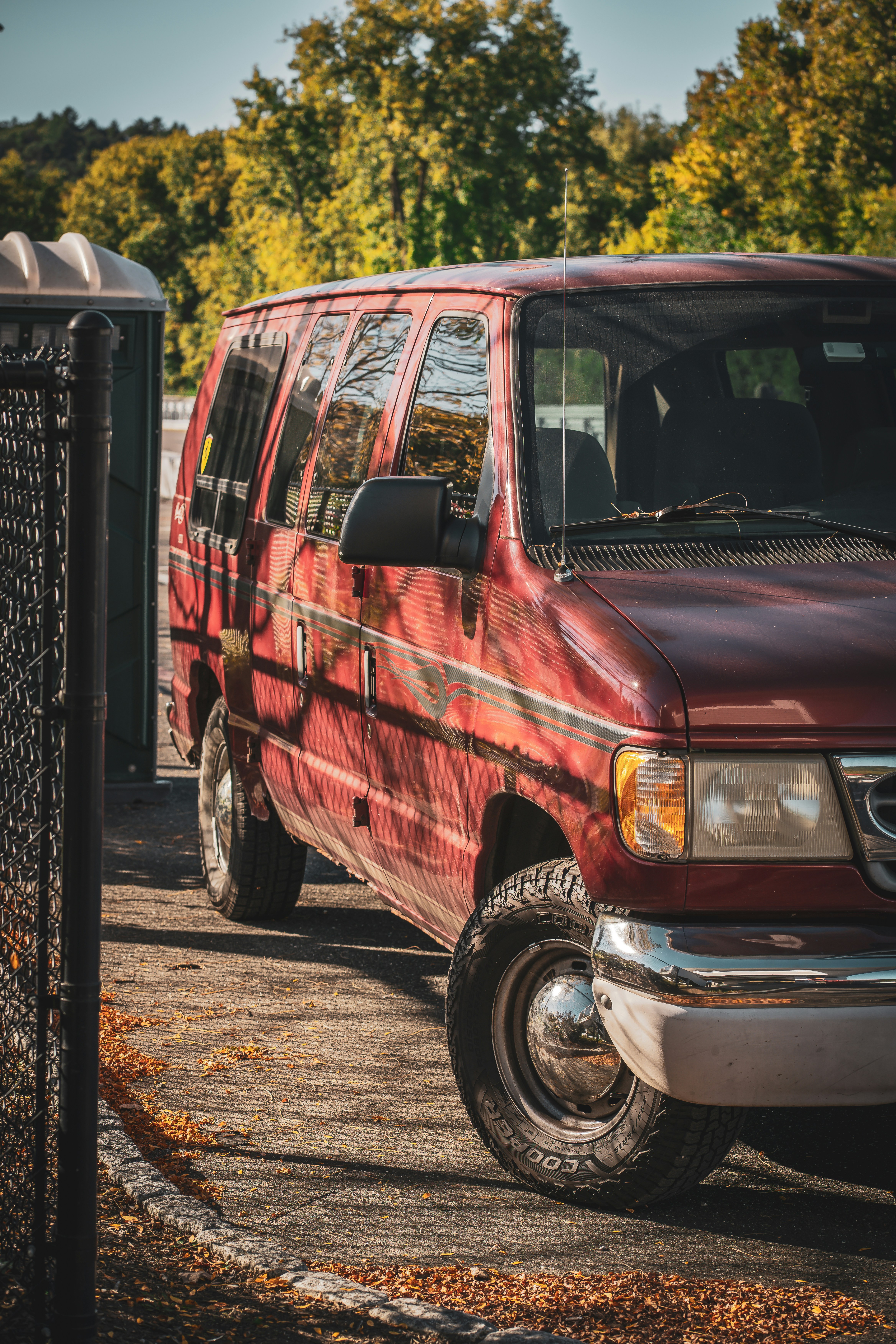 Red van parked beside a fence, surrounded by autumn foliage and soft sunlight. The scene captures a moment of stillness in a vibrant outdoor setting.