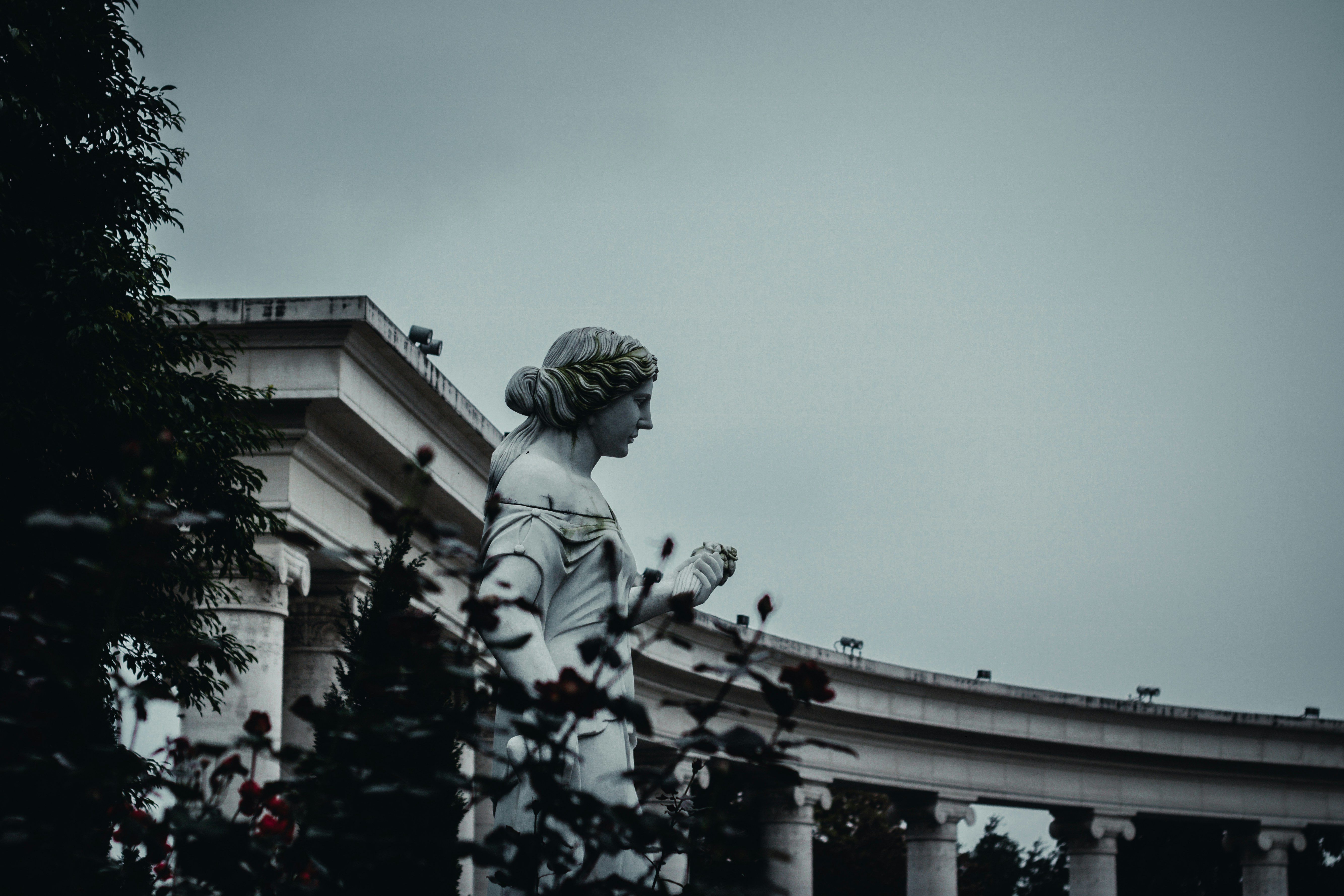A marble statue of a woman stands amidst blooming roses, framed by a classical colonnade under a moody sky.