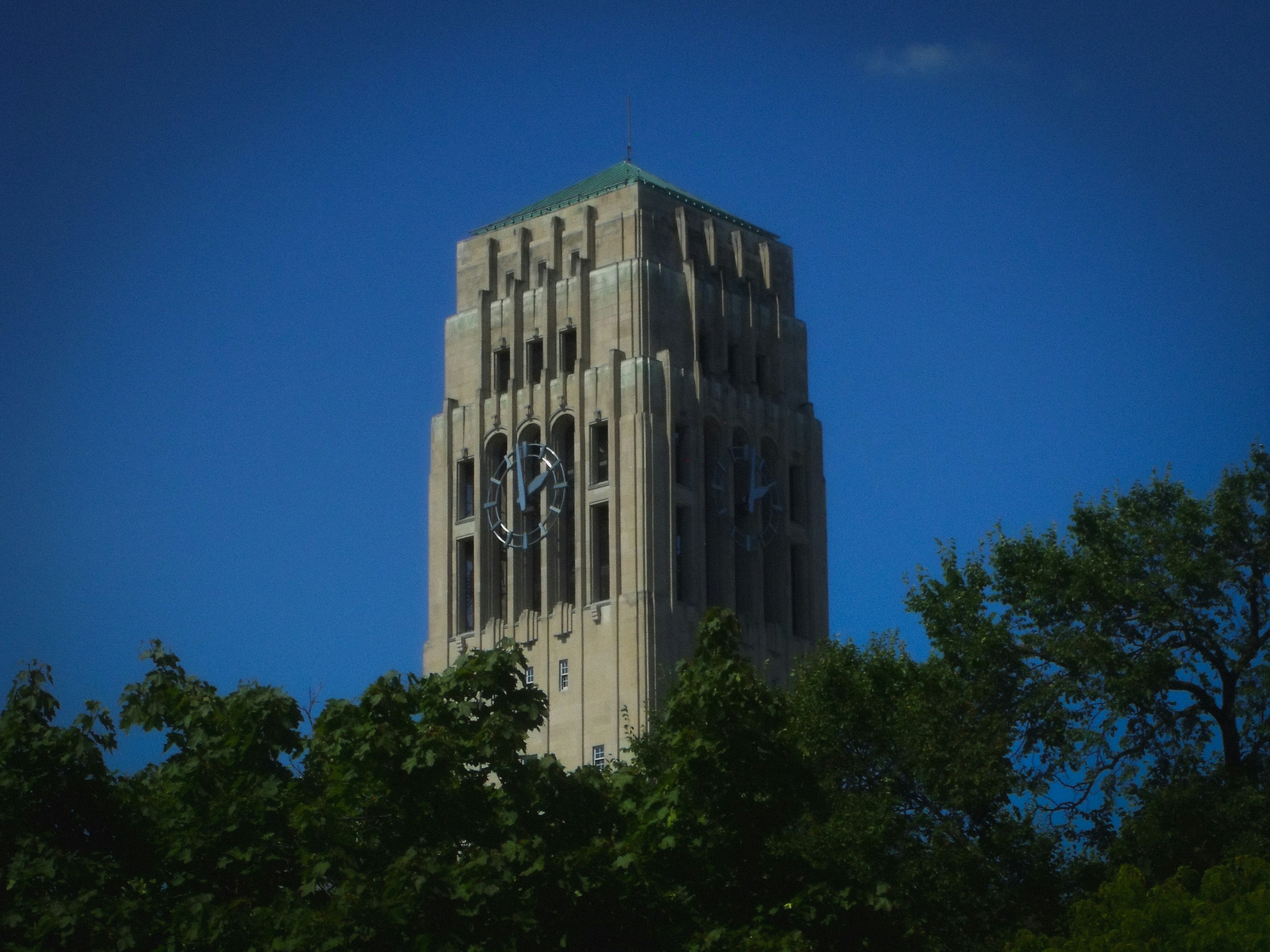 Art Deco clock tower rising above lush greenery against a clear blue sky.