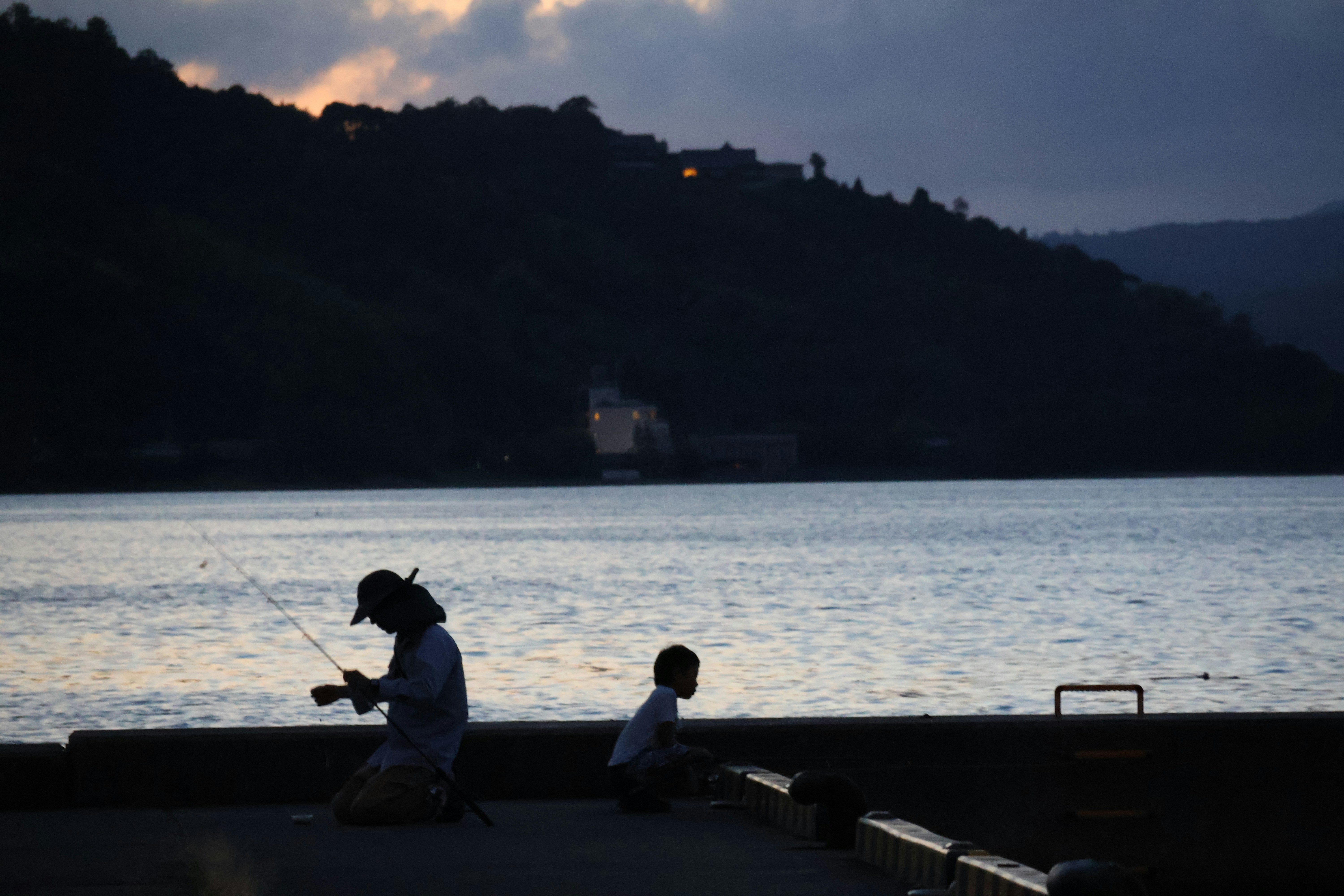 Two figures fishing by the water at dusk.