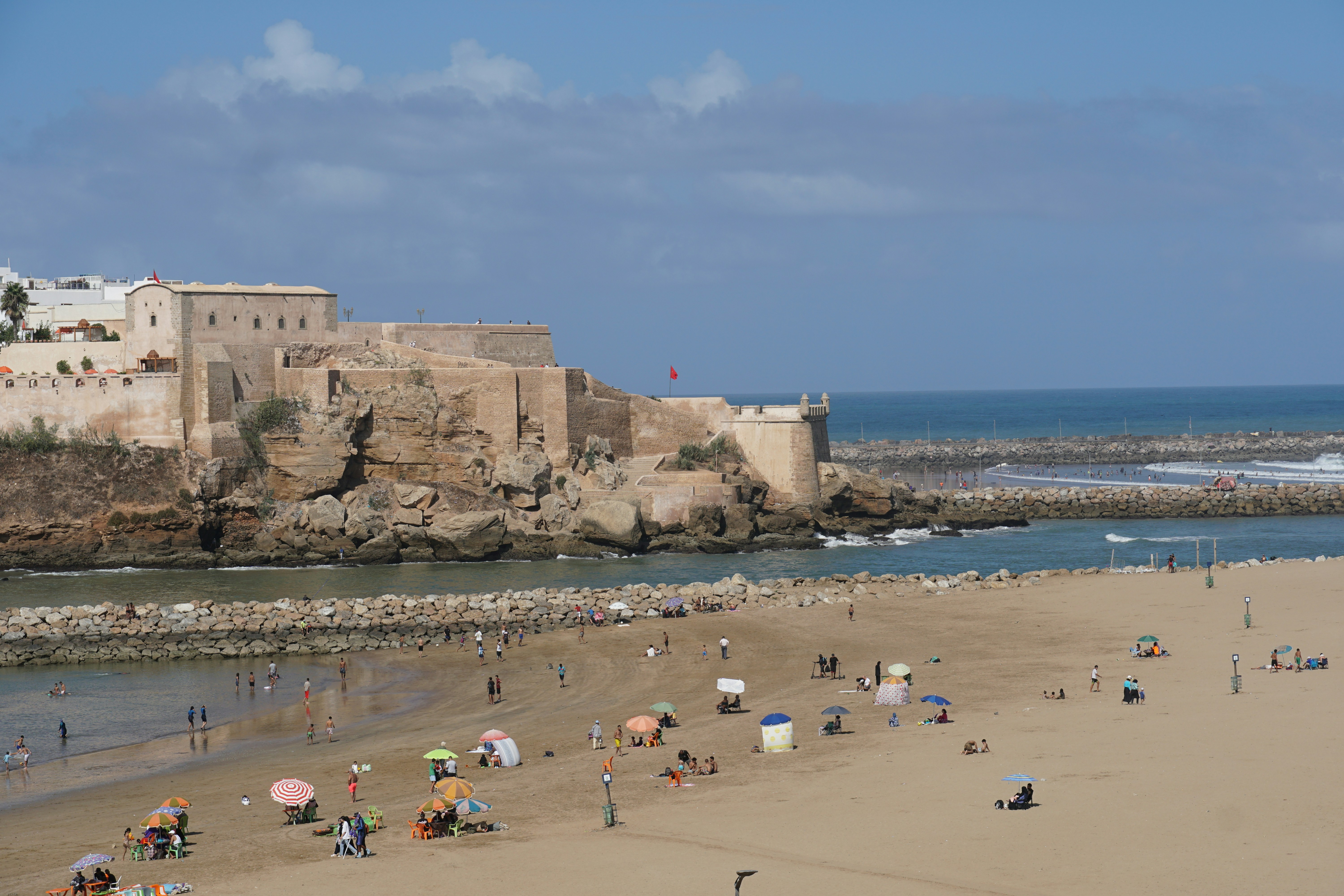 Historic coastal fortification overlooking a sandy beach filled with sunbathers and colorful umbrellas. Gentle waves lap at the shore.
