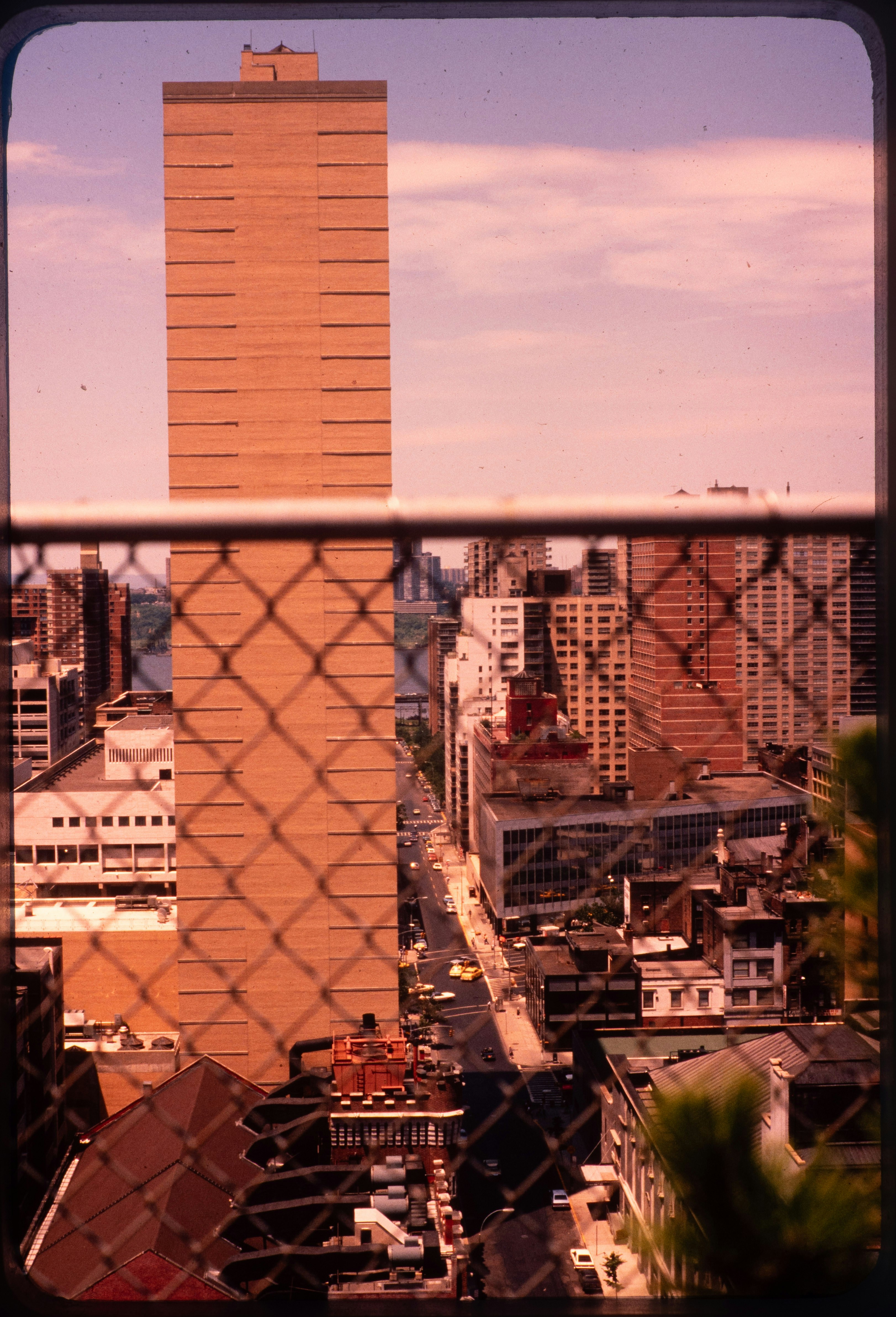 A tall, beige skyscraper dominates the city skyline, framed by a fence, with a view of bustling streets below.