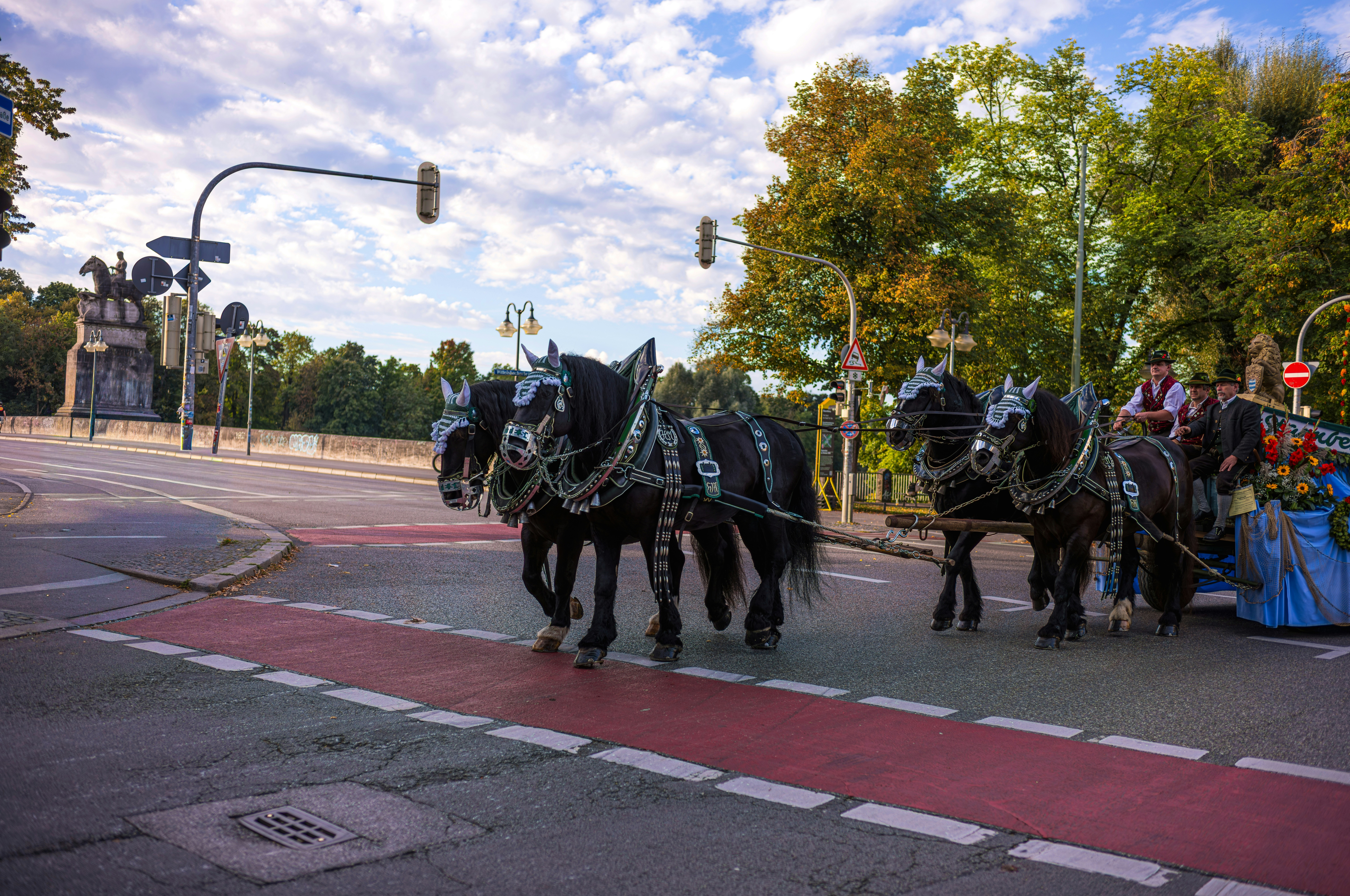 Horse-drawn carriage traversing a scenic road, adorned with decorative harnesses and colorful floral arrangements, set against a backdrop of lush trees and a cloudy sky.