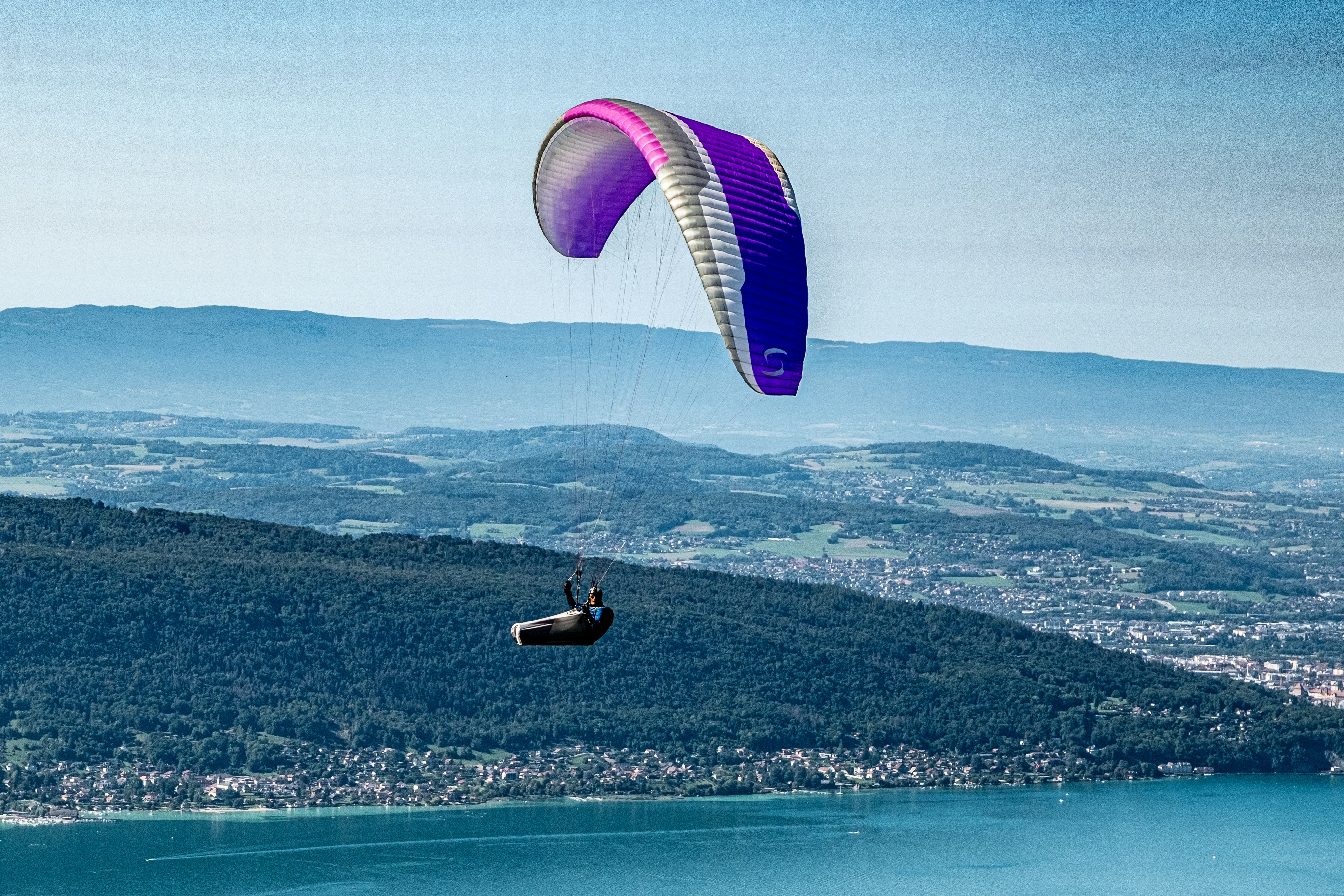 A solo paraglider rides the thermals at Col de la Forclaz, in the Aravis mountains, with the town and lake of Annecy in the background | Person paragliding over a lake and forested hills.