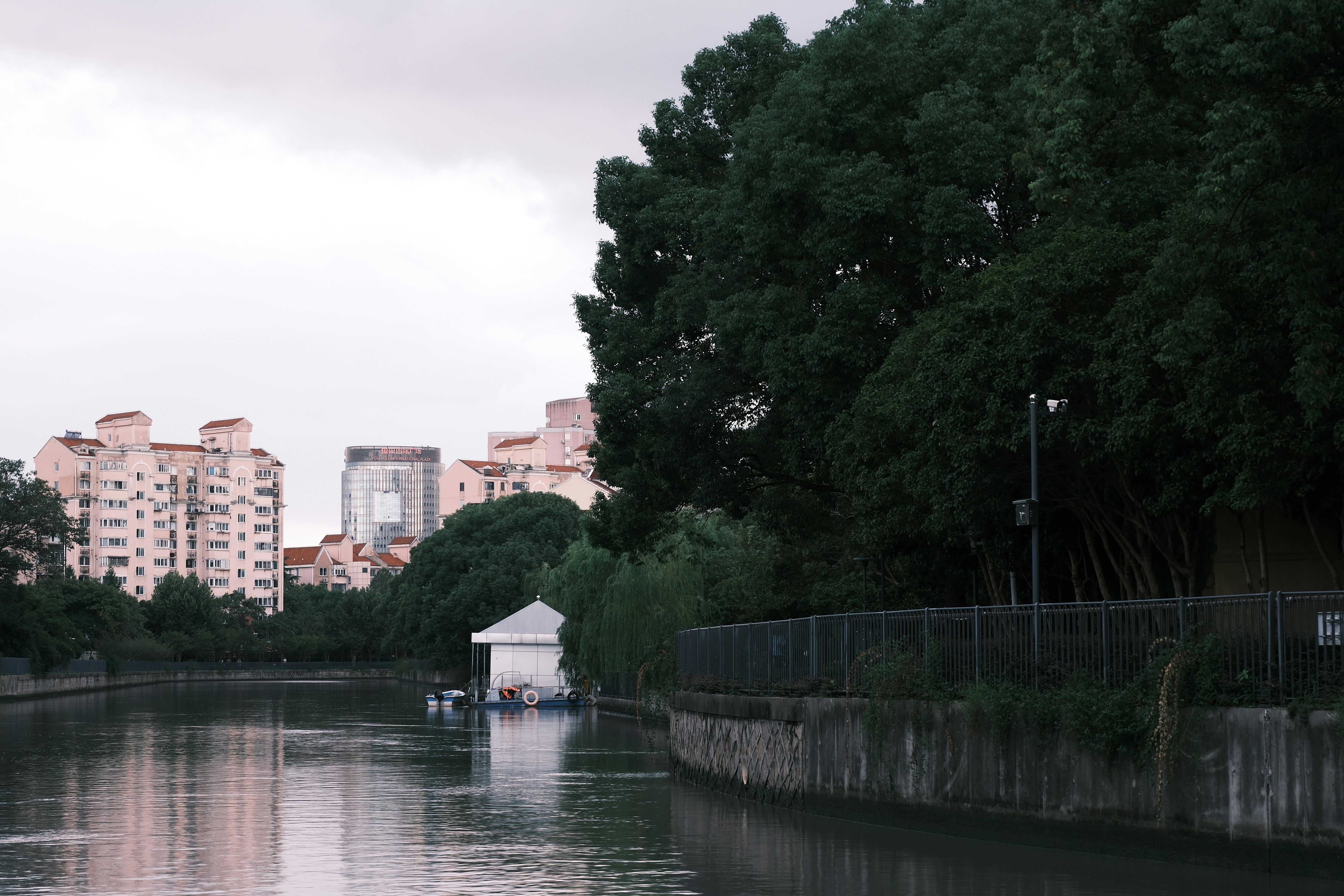 A serene urban canal flanked by dense green trees, with a quiet city skyline in the background | River flowing past buildings and trees under cloudy sky.