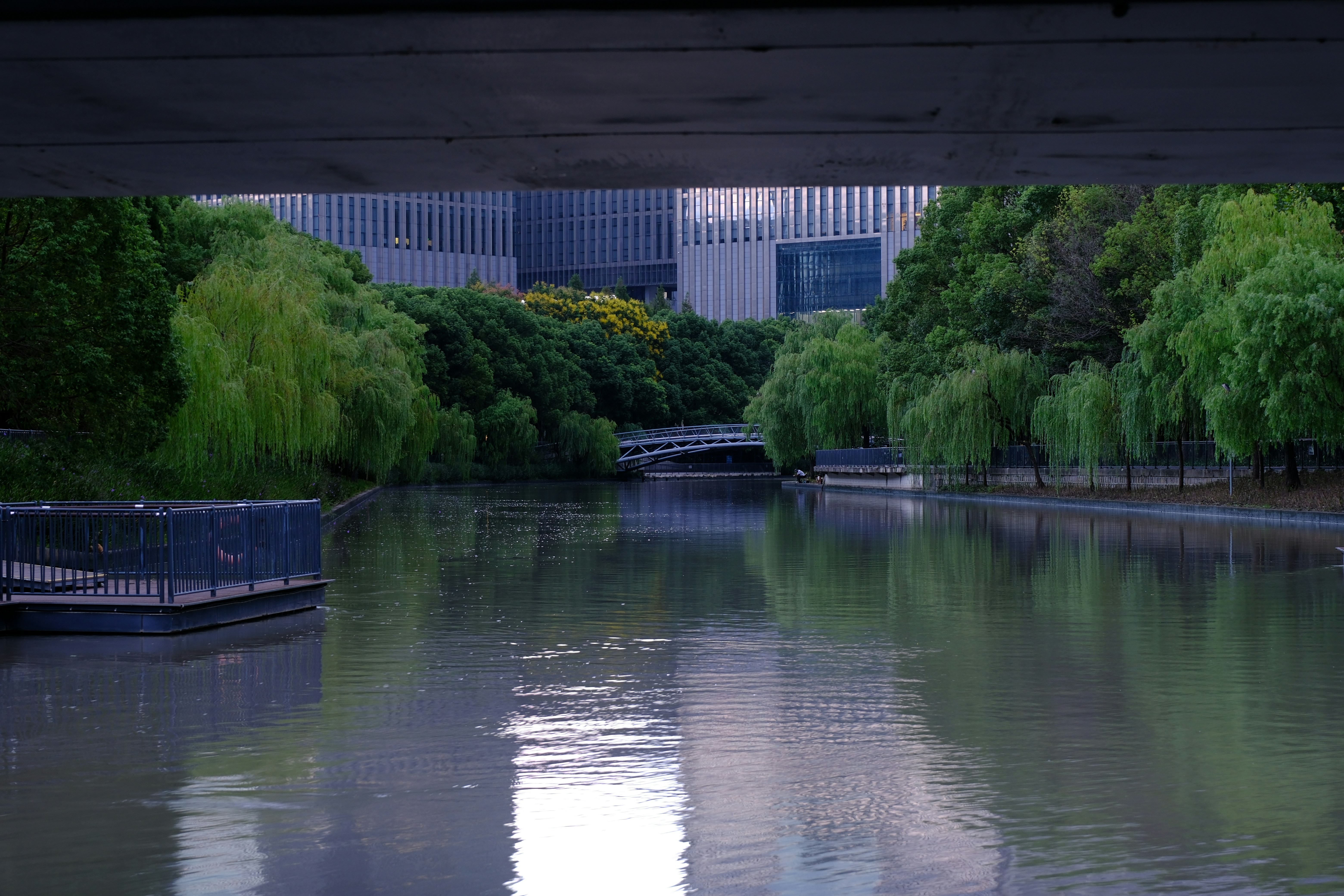 A cinematic frame from under a bridge, overlooking a calm canal, a floating dock, and distant city buildings softened by nature | Calm river reflects modern buildings surrounded by lush trees.