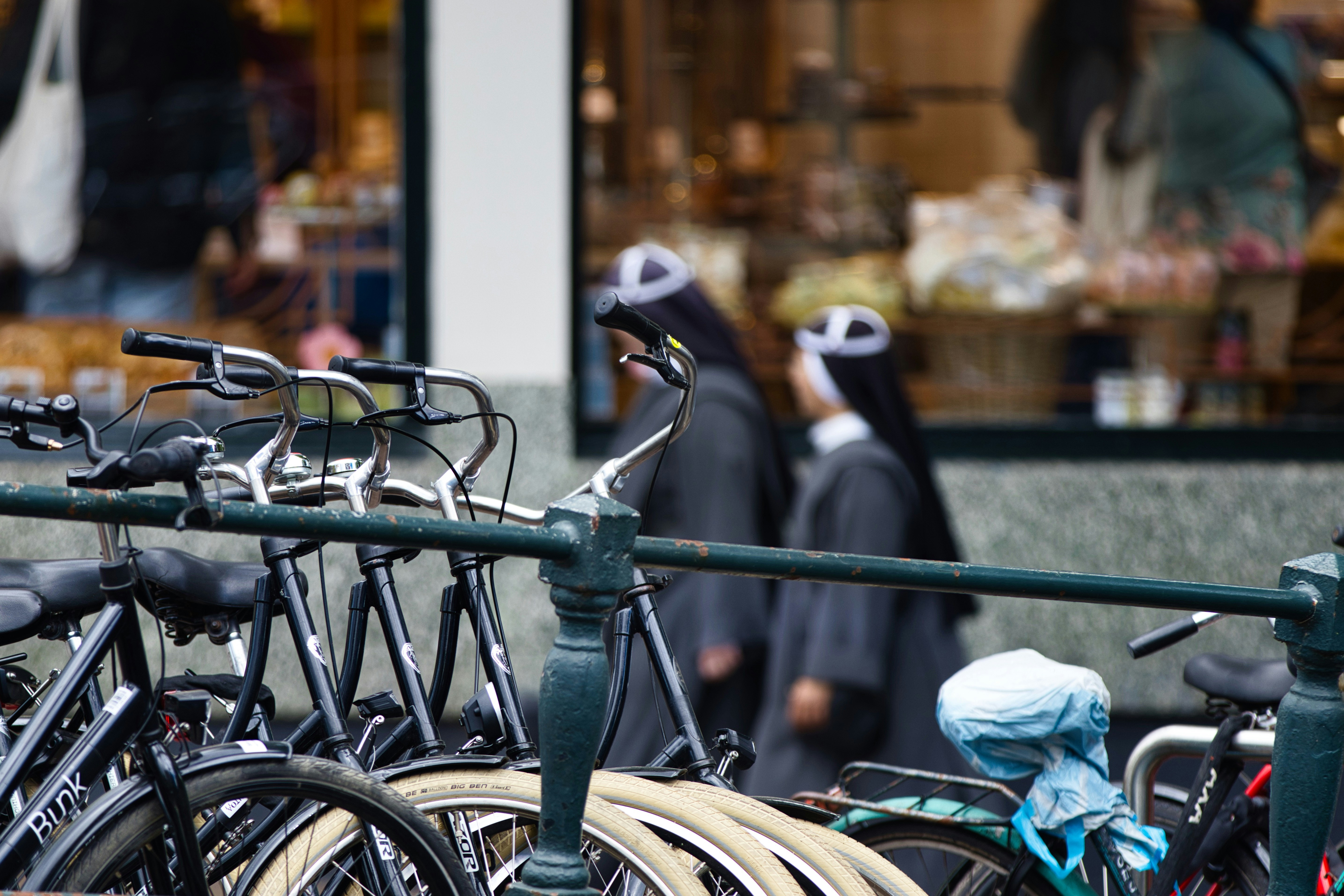 Two nuns walk past a row of bicycles in a bustling urban setting, highlighting a blend of tradition and modern life. The scene captures a quiet moment in a busy environment.