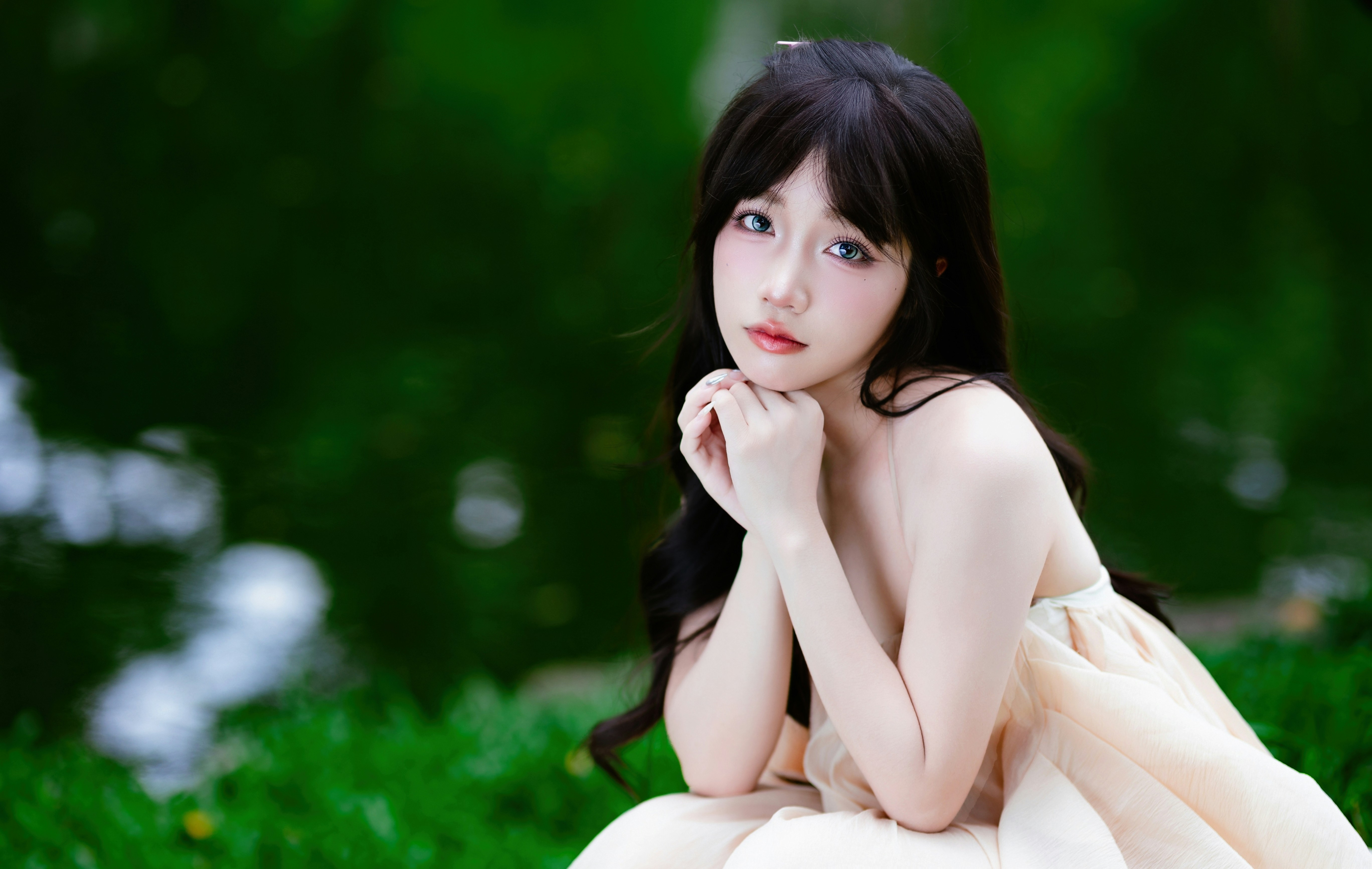 A young woman in a soft, flowing dress gazes thoughtfully while seated on lush grass beside a tranquil water surface.
