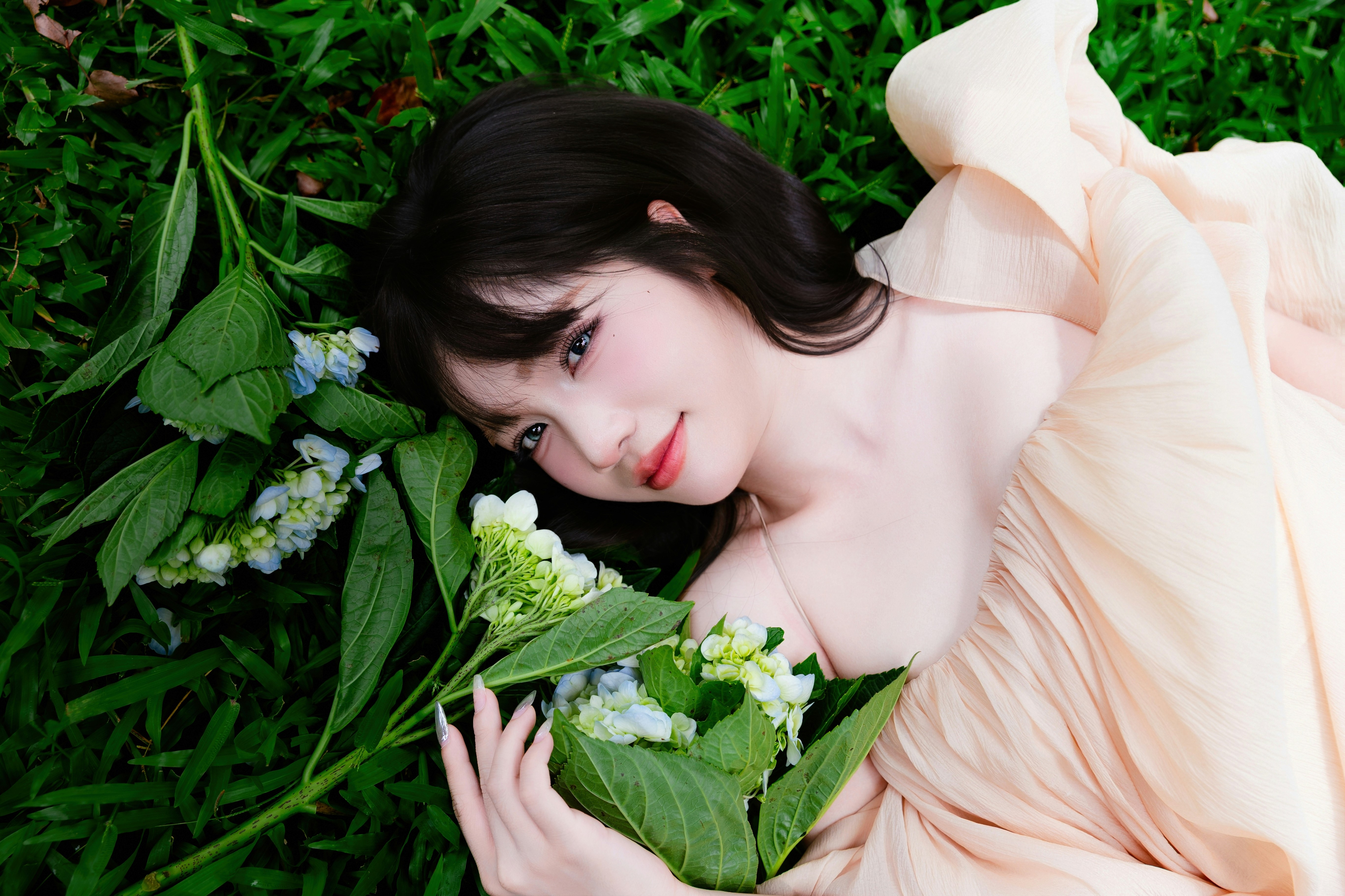 Smiling young woman lying on green grass with a bouquet of fresh hydrangea flowers, wearing a soft cream dress in natural light. | Young woman in flowing dress lying on grass with flowers.