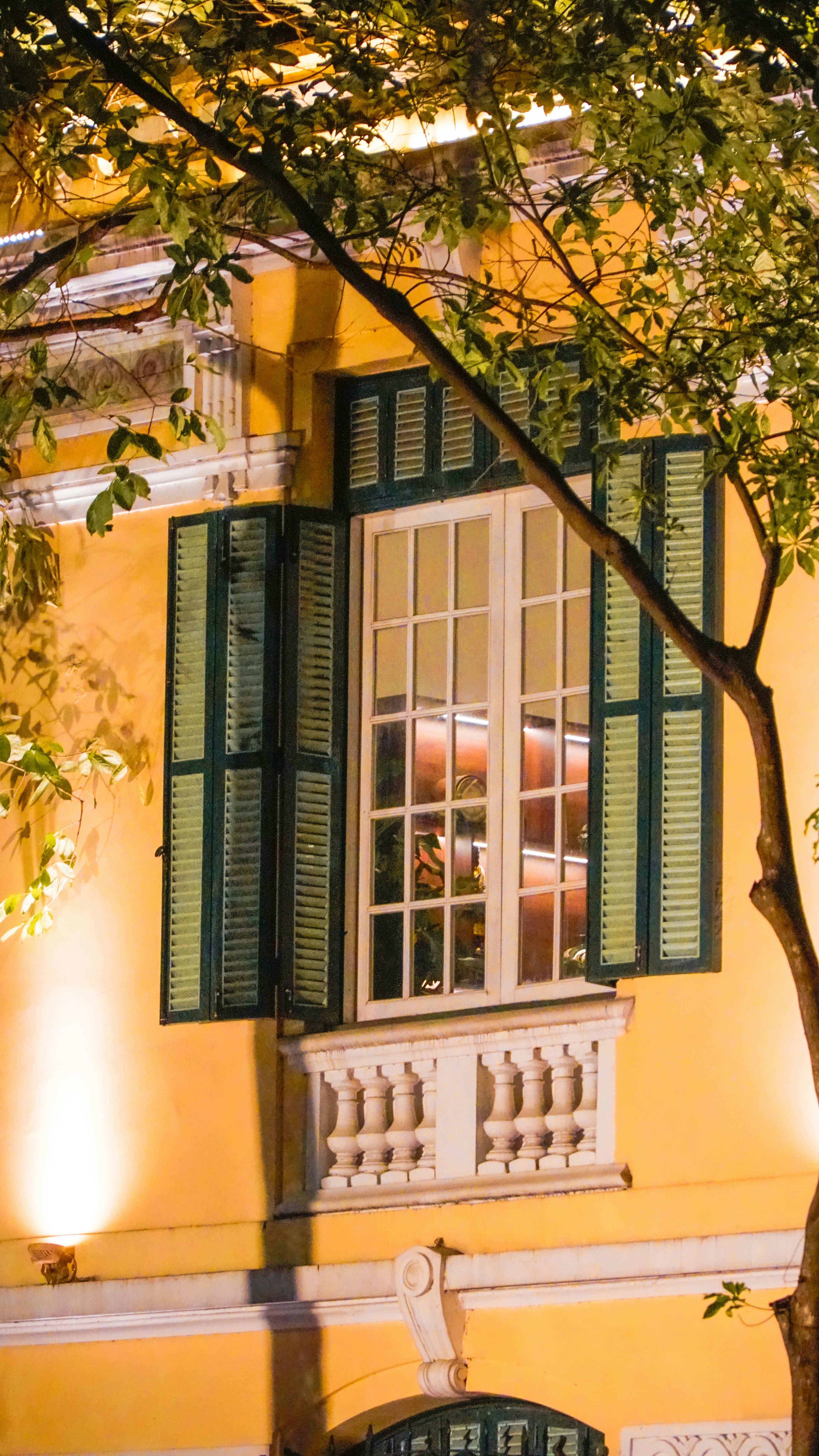 Nighttime photograph of a warm yellow building with open emerald shutters and a white balcony. A softly lit window glows through the opening, framed by overhanging tree branches.