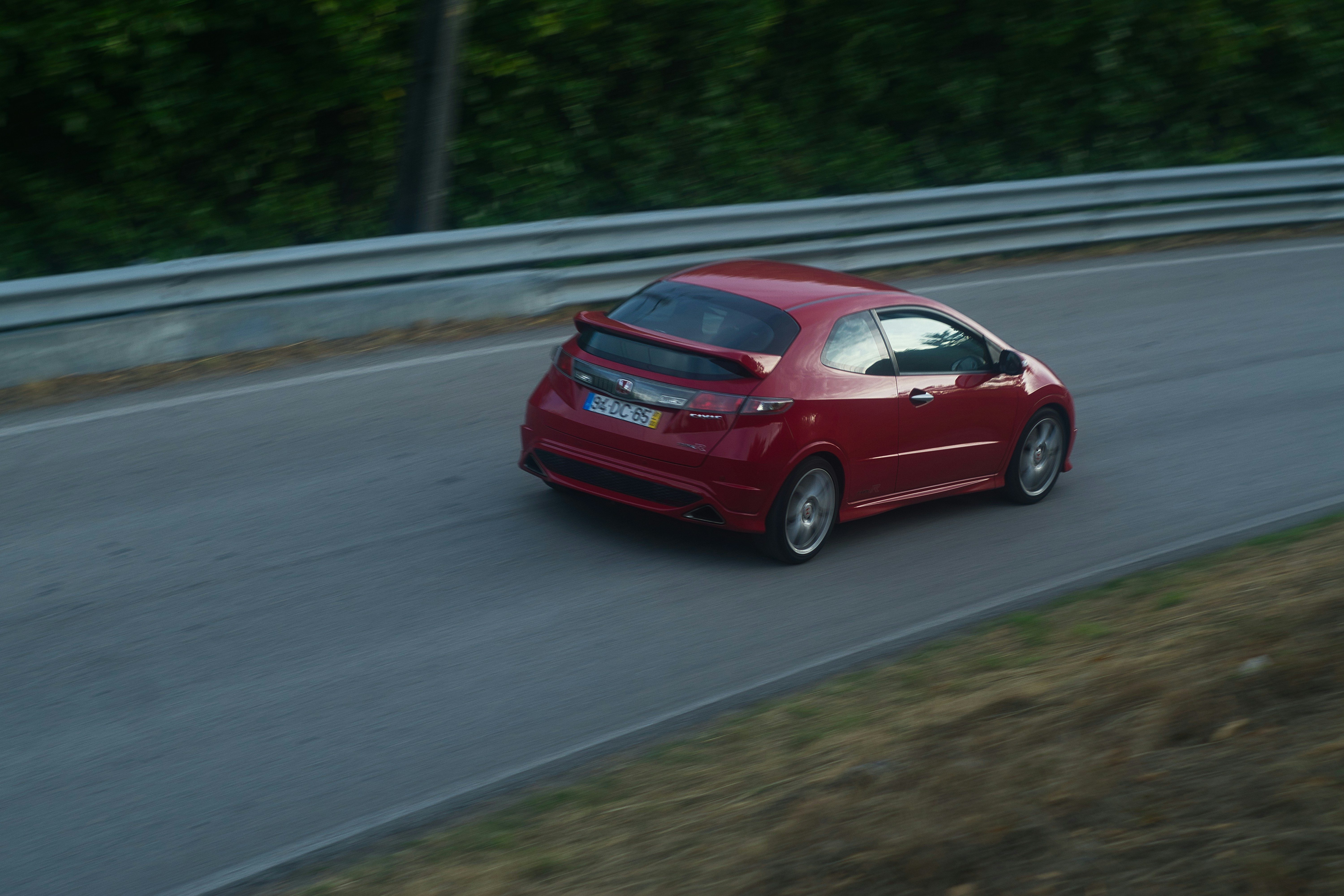 A red hatchback car speeds around a curve on a winding road, surrounded by lush greenery.