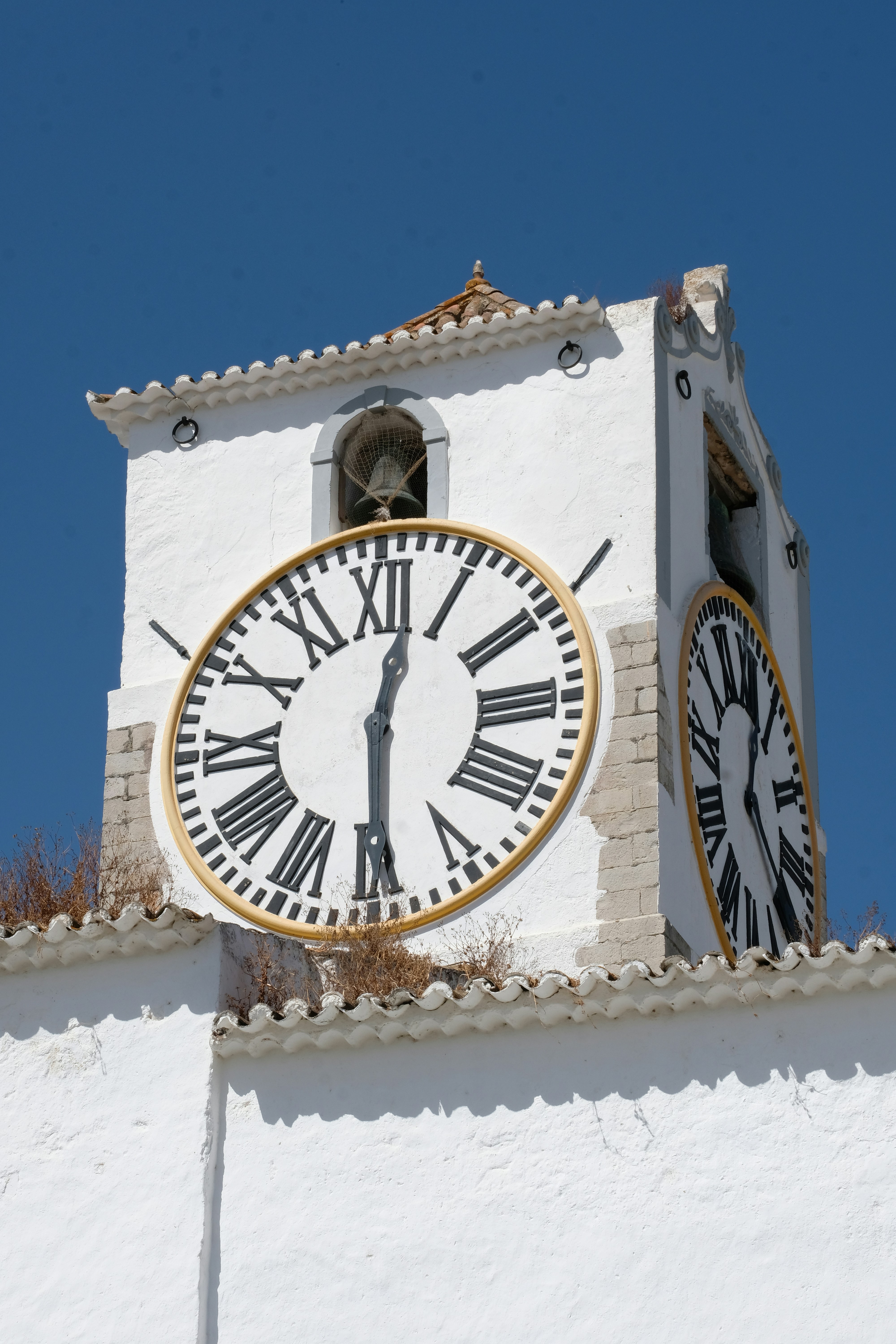 Clock tower with large Roman numeral faces in Tavira, Spain, under clear blue sky. | White clock tower with roman numerals on face