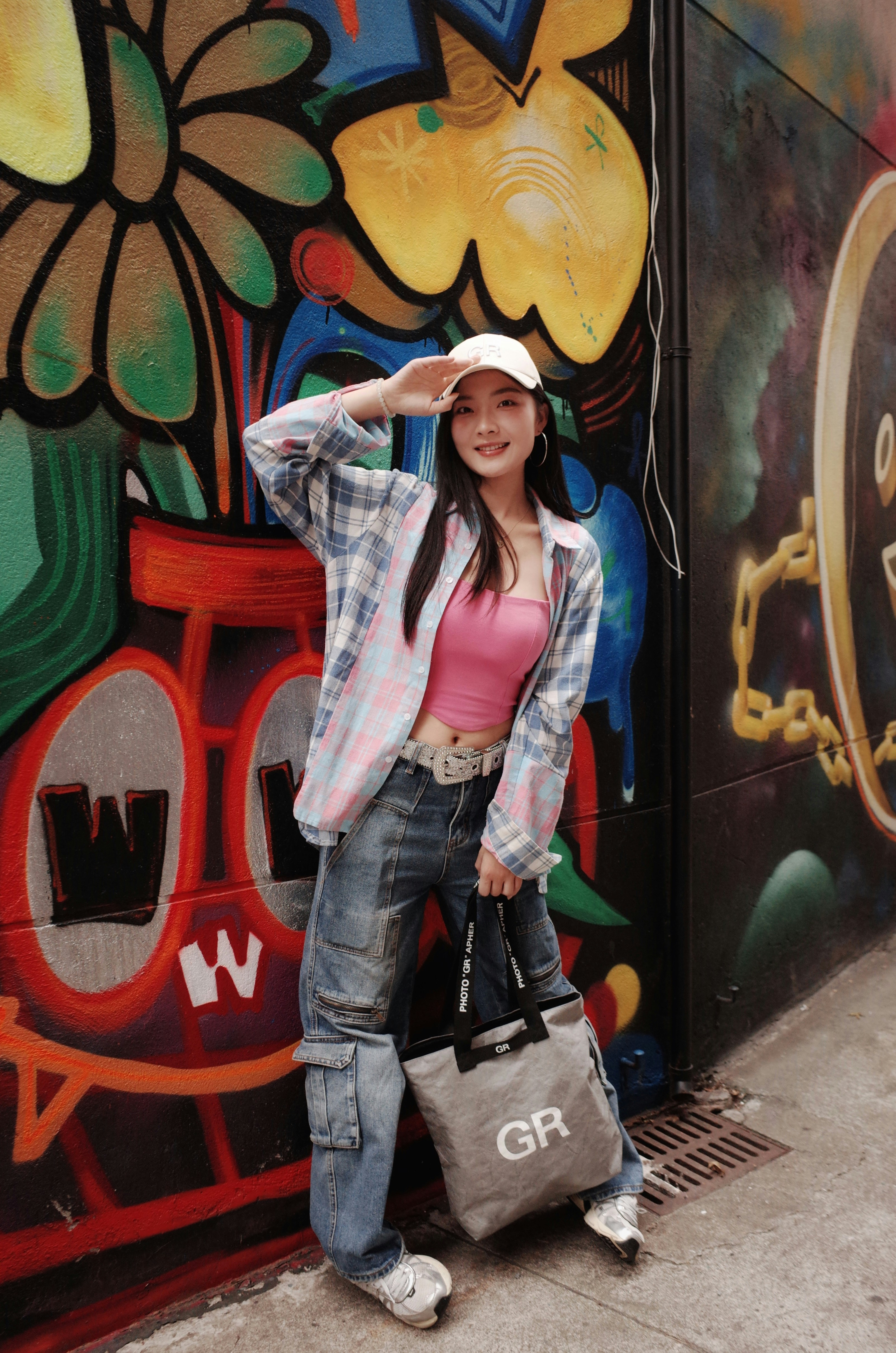 Young woman posing against a vibrant graffiti wall, showcasing a casual streetwear style with a playful salute. Bright colors and artistic elements frame the scene.