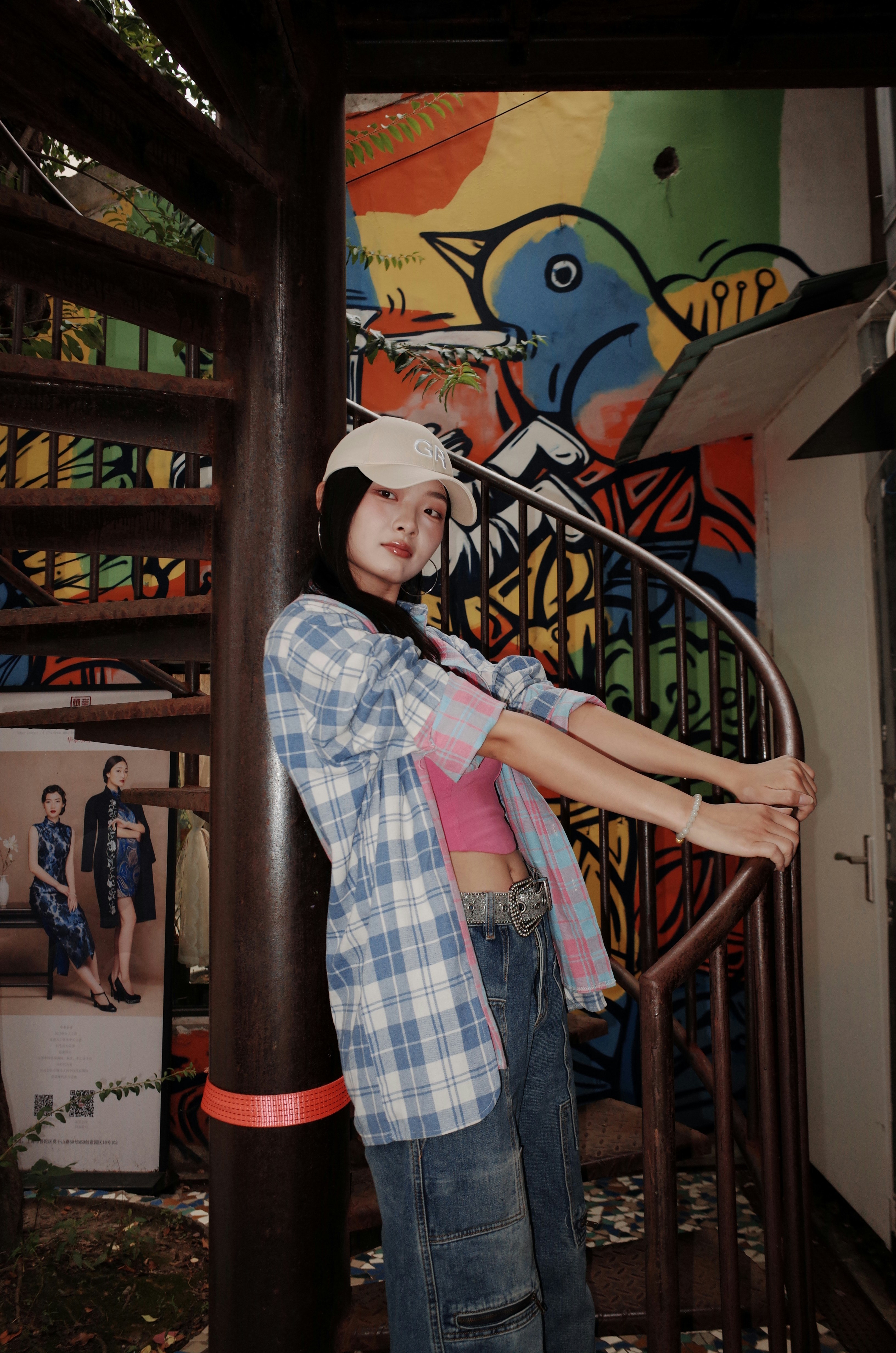 A young woman poses gracefully on a spiral staircase adorned with vibrant street art. Her casual outfit and expressive pose complement the lively background.