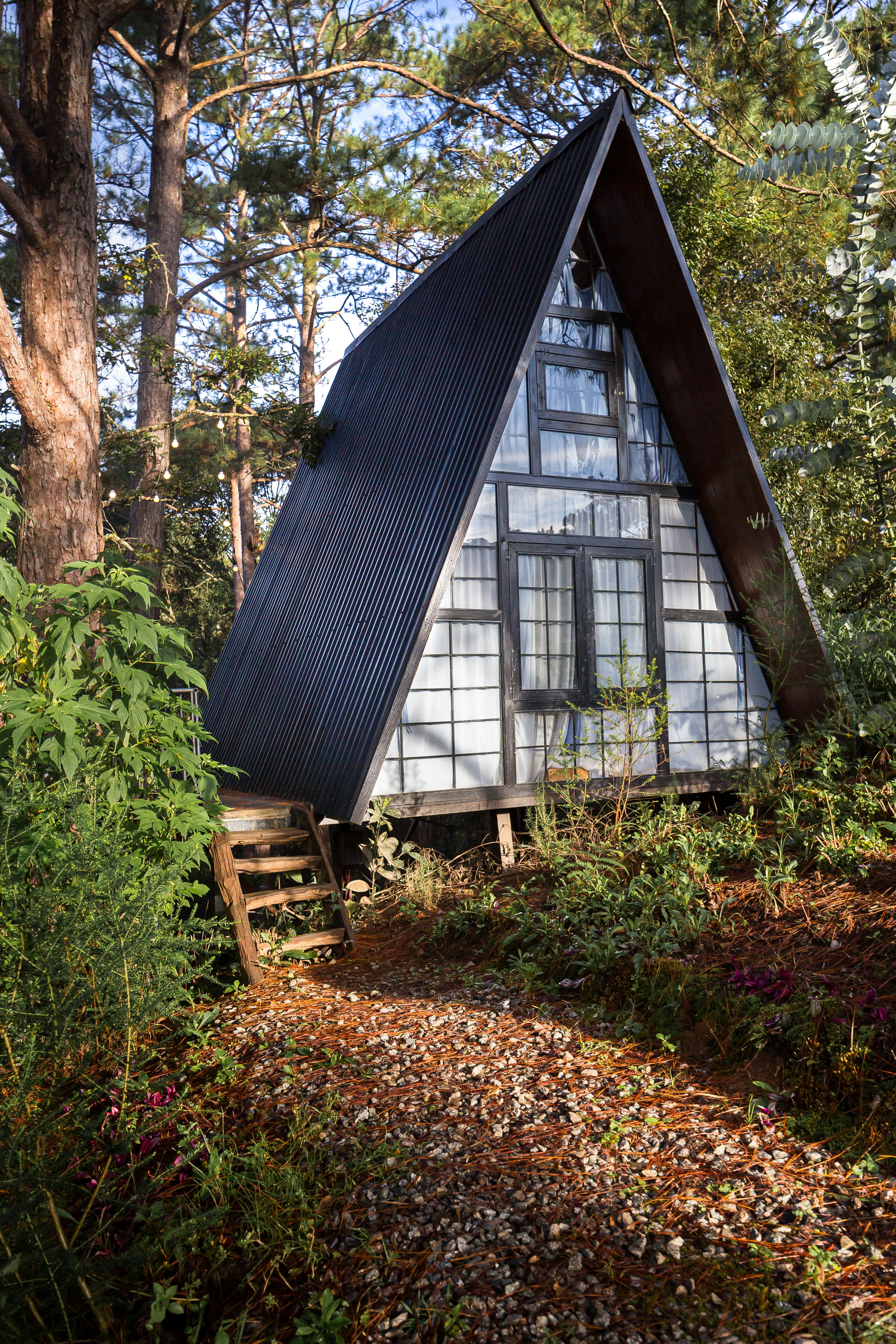 Charming A-frame cabin nestled among lush greenery, with a stone pathway leading to its entrance. The cabin's unique architecture harmonizes with the surrounding forest.
