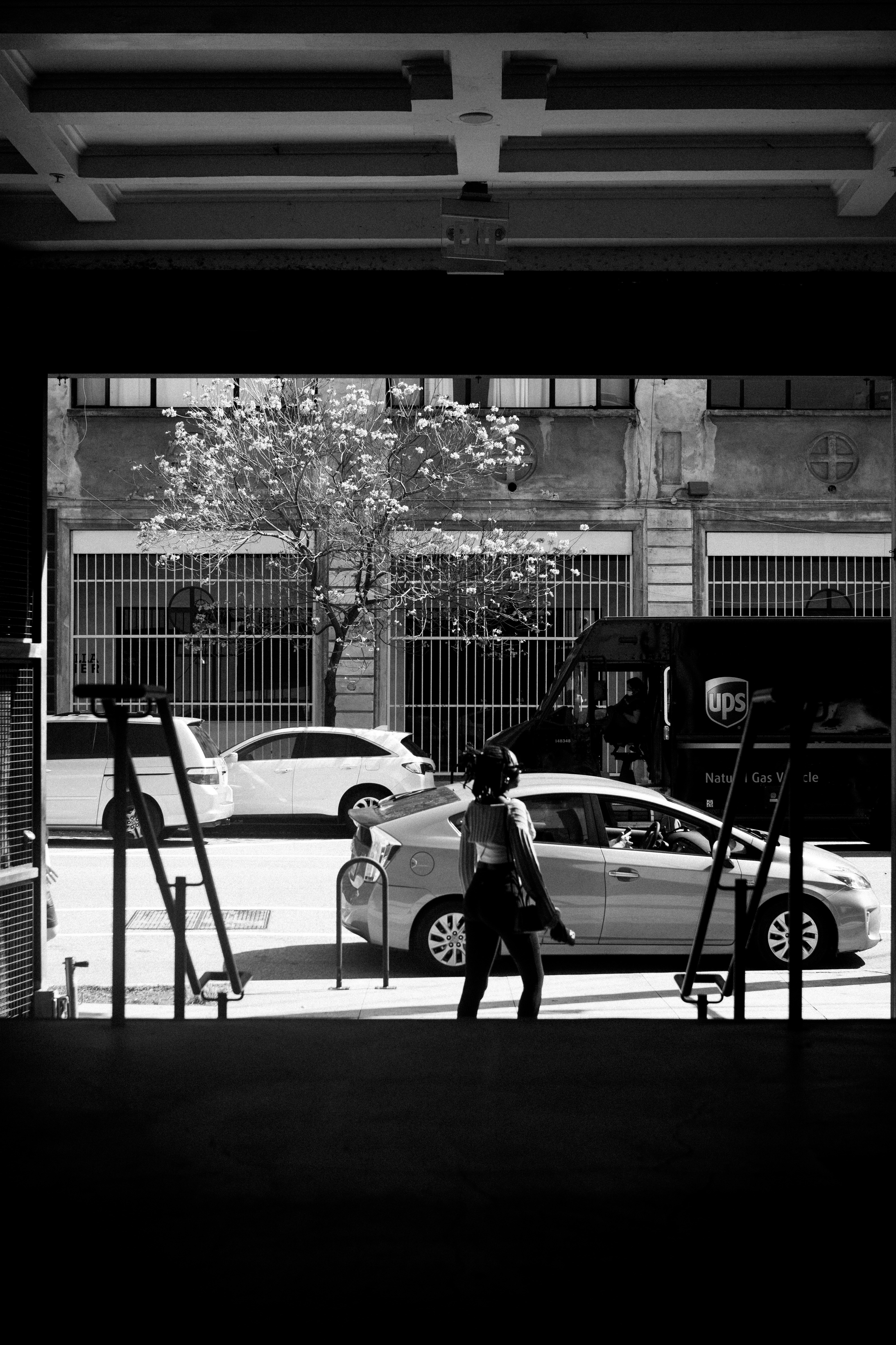 A woman walks past parked cars and a delivery truck, framed by the architectural details of a building entrance. The scene is presented in black and white, emphasizing contrasts and urban textures.