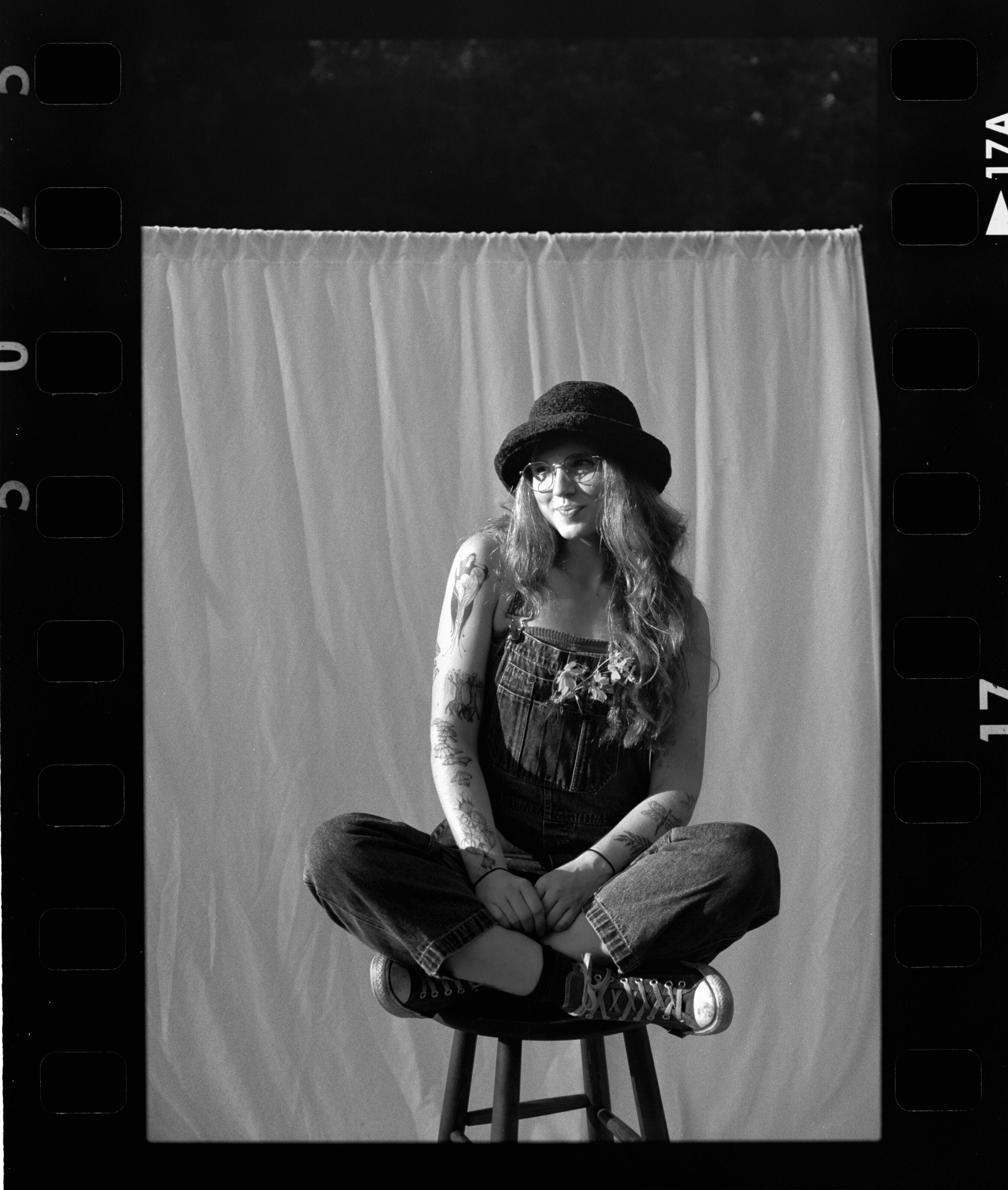 A young woman with tattoos and glasses sits cross-legged on a stool, wearing overalls and a hat, against a softly draped background.