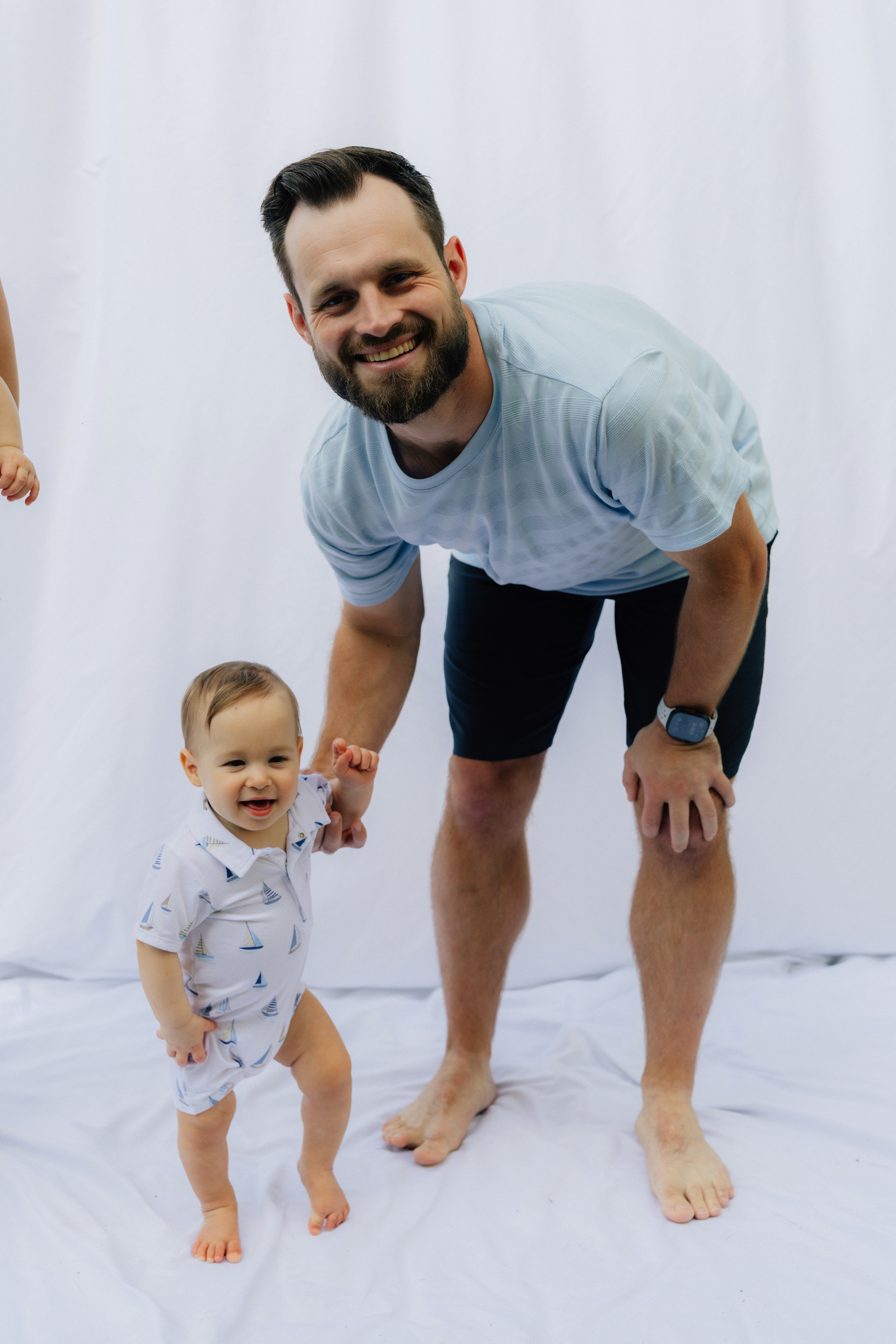 A cheerful toddler in a sailboat-patterned onesie holds hands with a smiling adult against a soft white backdrop.