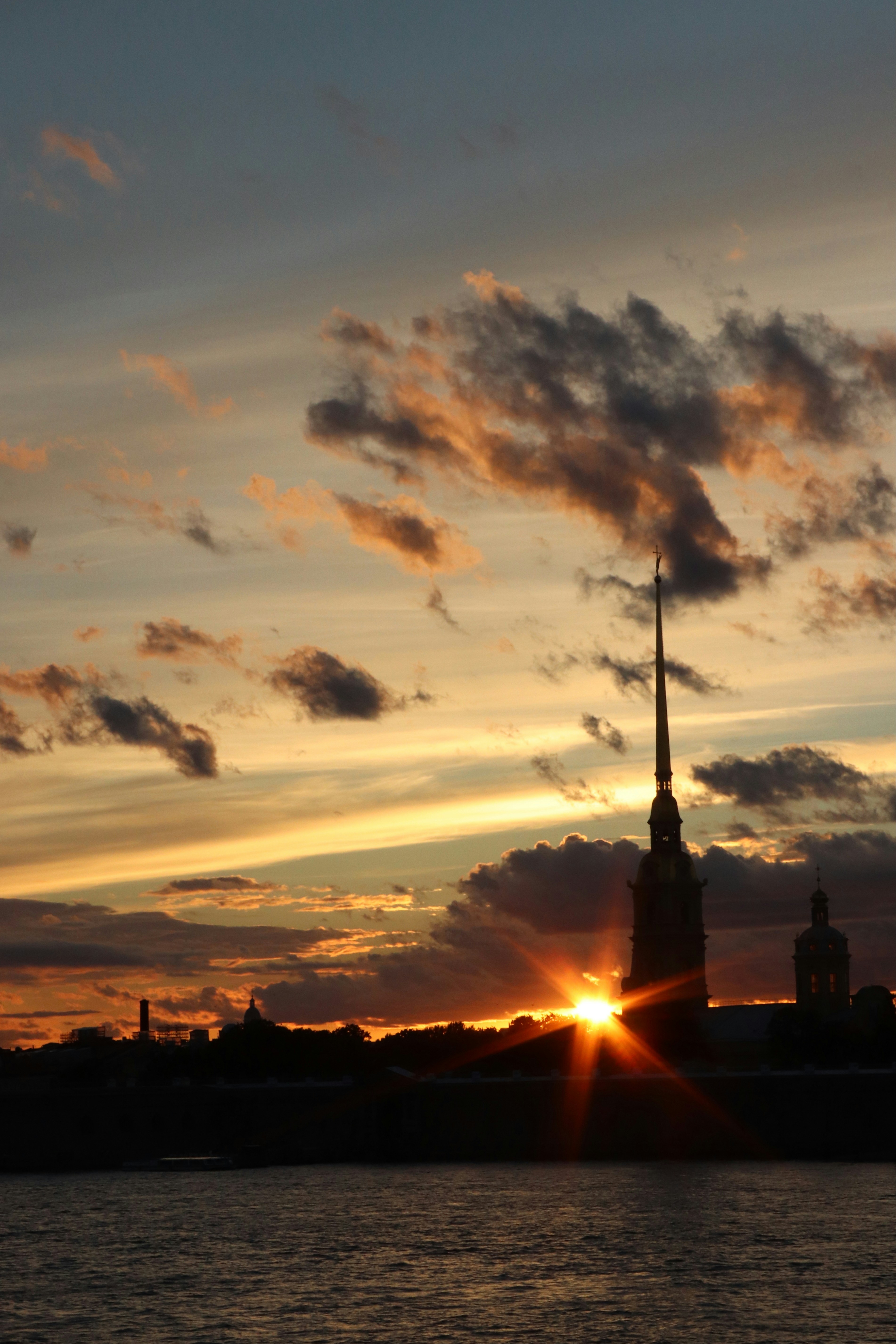 The silhouette of a historic spire and buildings stands against a vibrant sunset, with clouds painted in warm hues. A tranquil waterway reflects the scene.