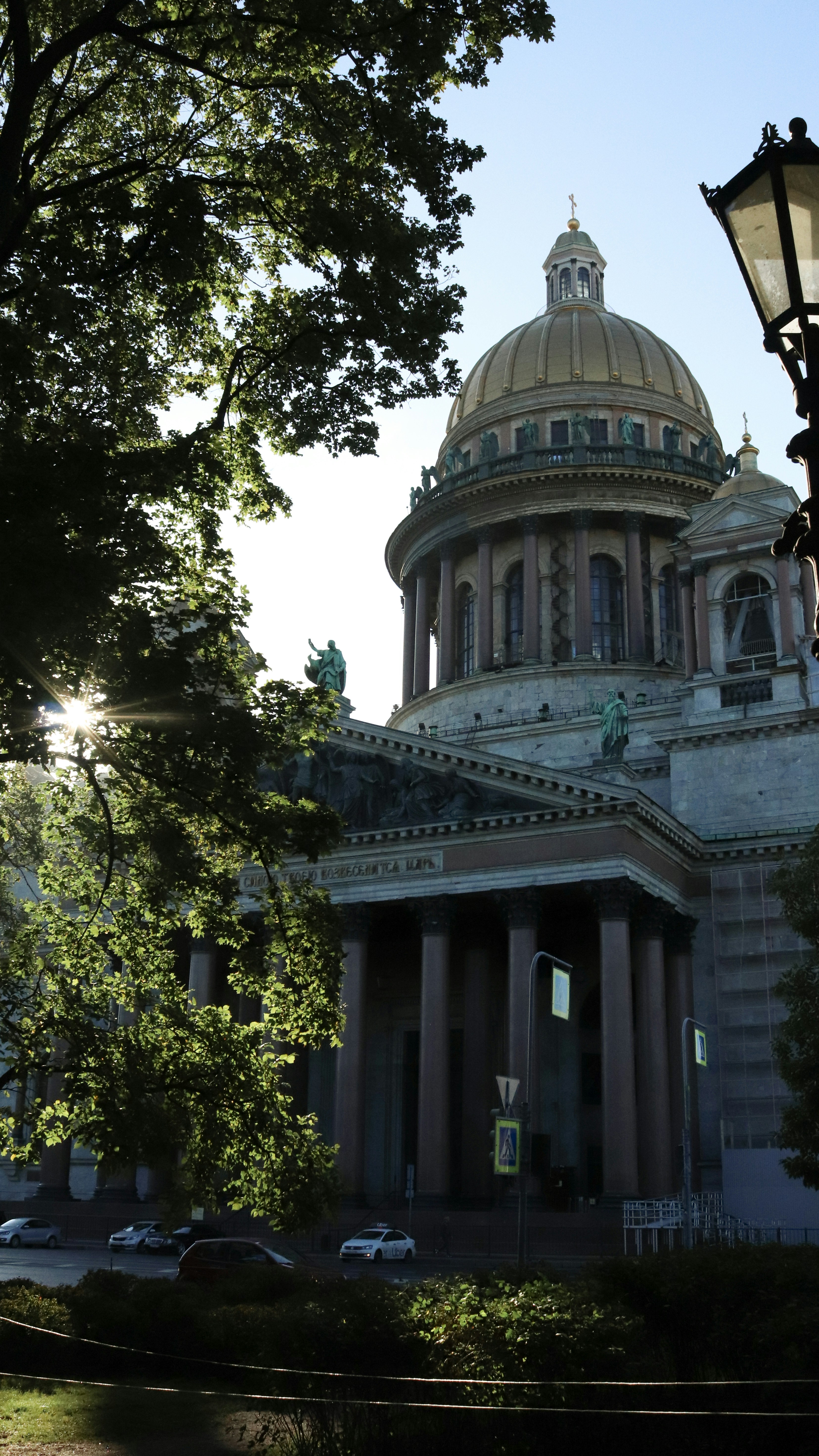 Historic cathedral with a grand dome and ornate sculptures, partially obscured by lush greenery. Sunlight glimmers through the trees, enhancing the scene.