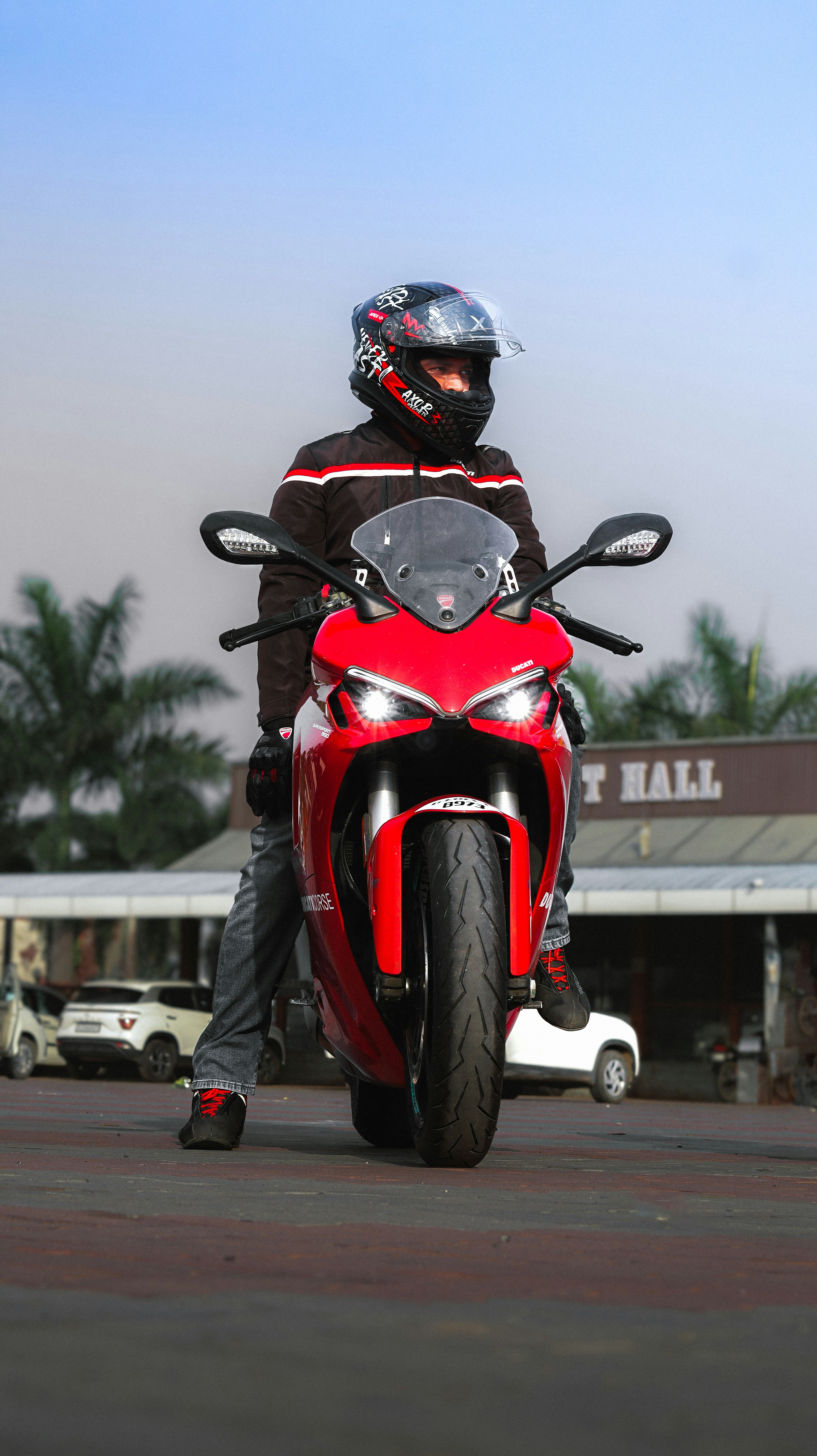 Motorcyclist in full gear confidently poses on a red sport bike, showcasing the sleek design against a blurred background of palm trees and a building.