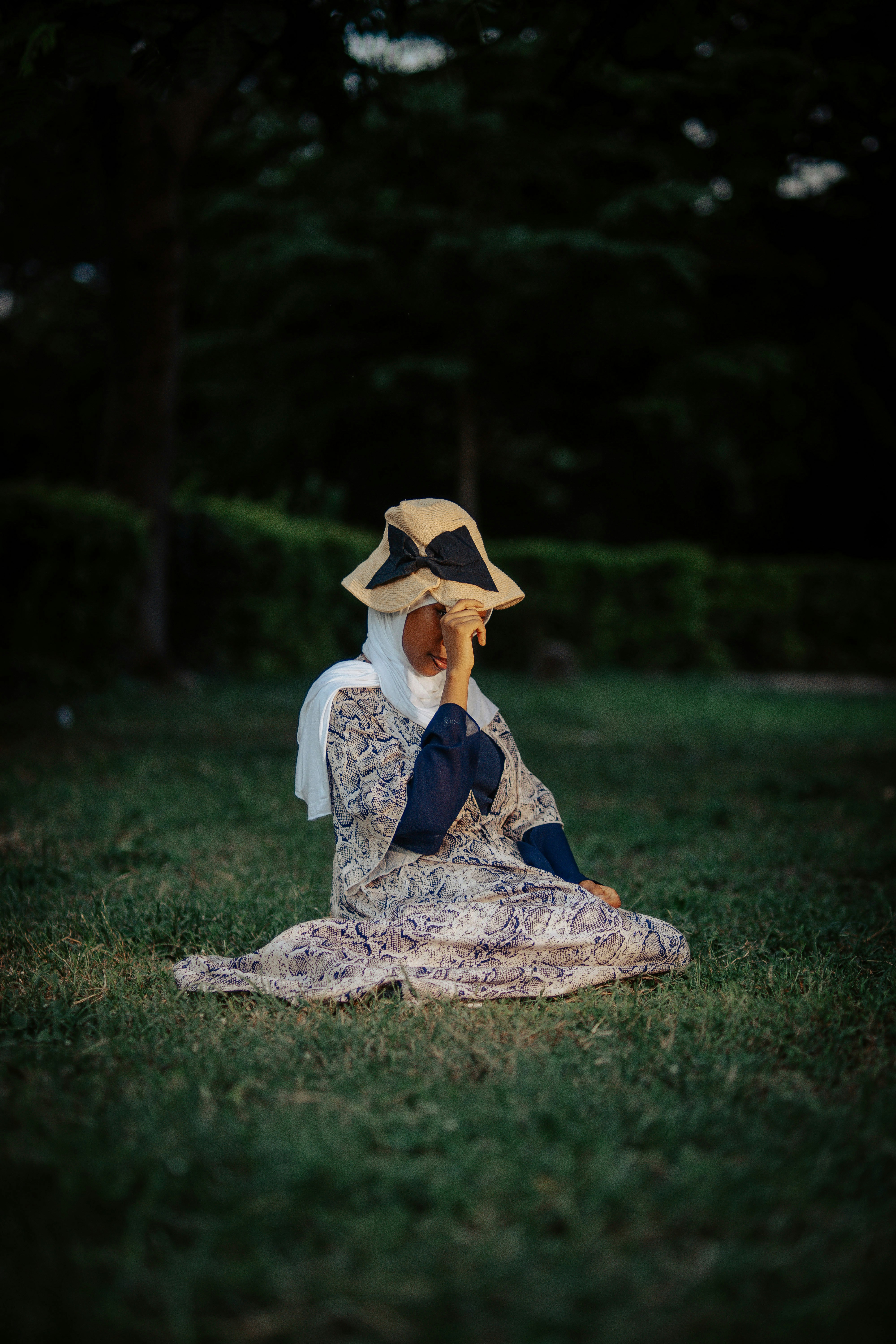 A young woman in a patterned dress and hat sits thoughtfully on the grass, embodying tranquility amidst a lush green backdrop.