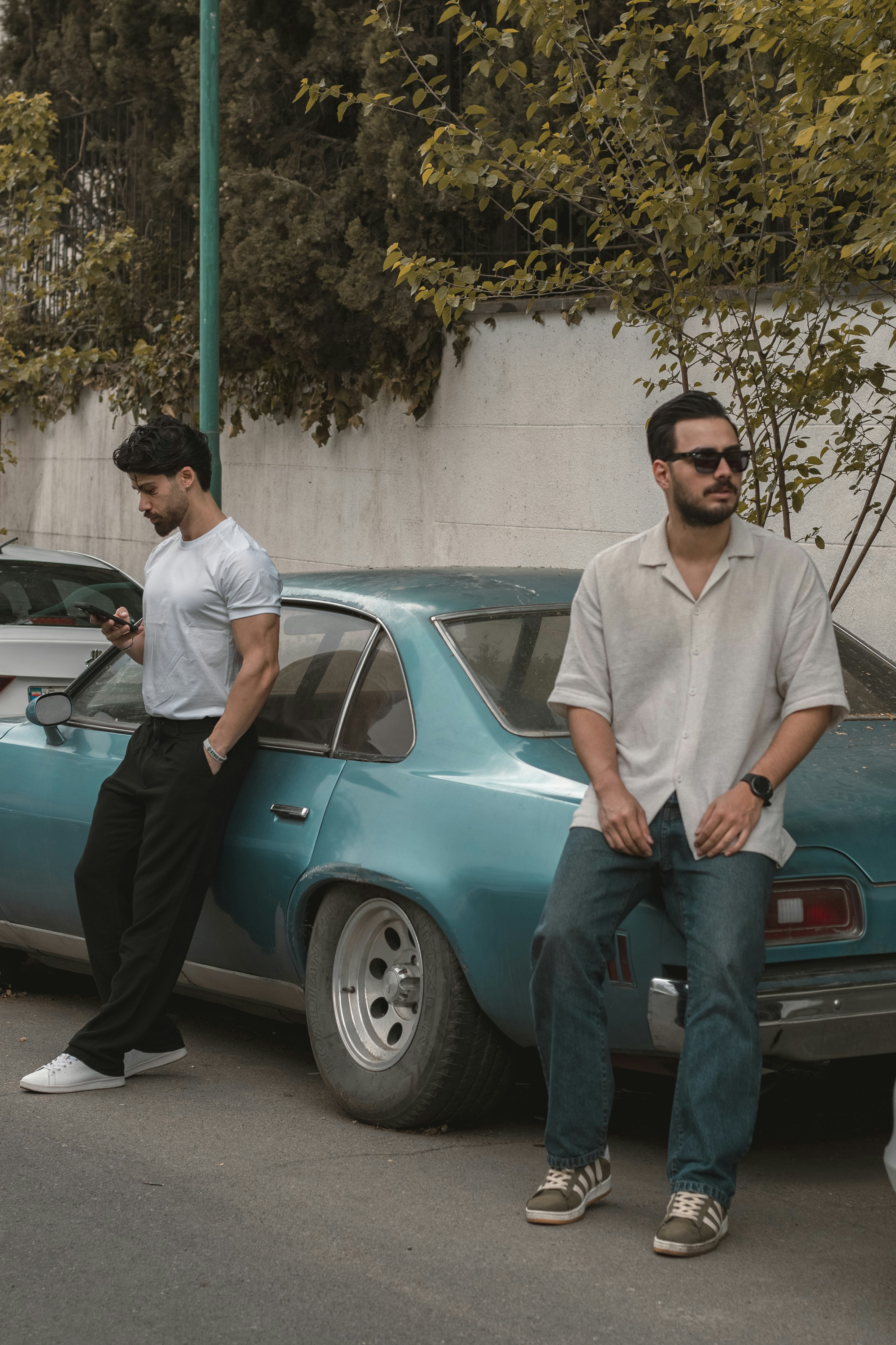 Two men casually leaning against a vintage car, one engrossed in his phone while the other observes his surroundings. The scene captures a relaxed urban atmosphere.