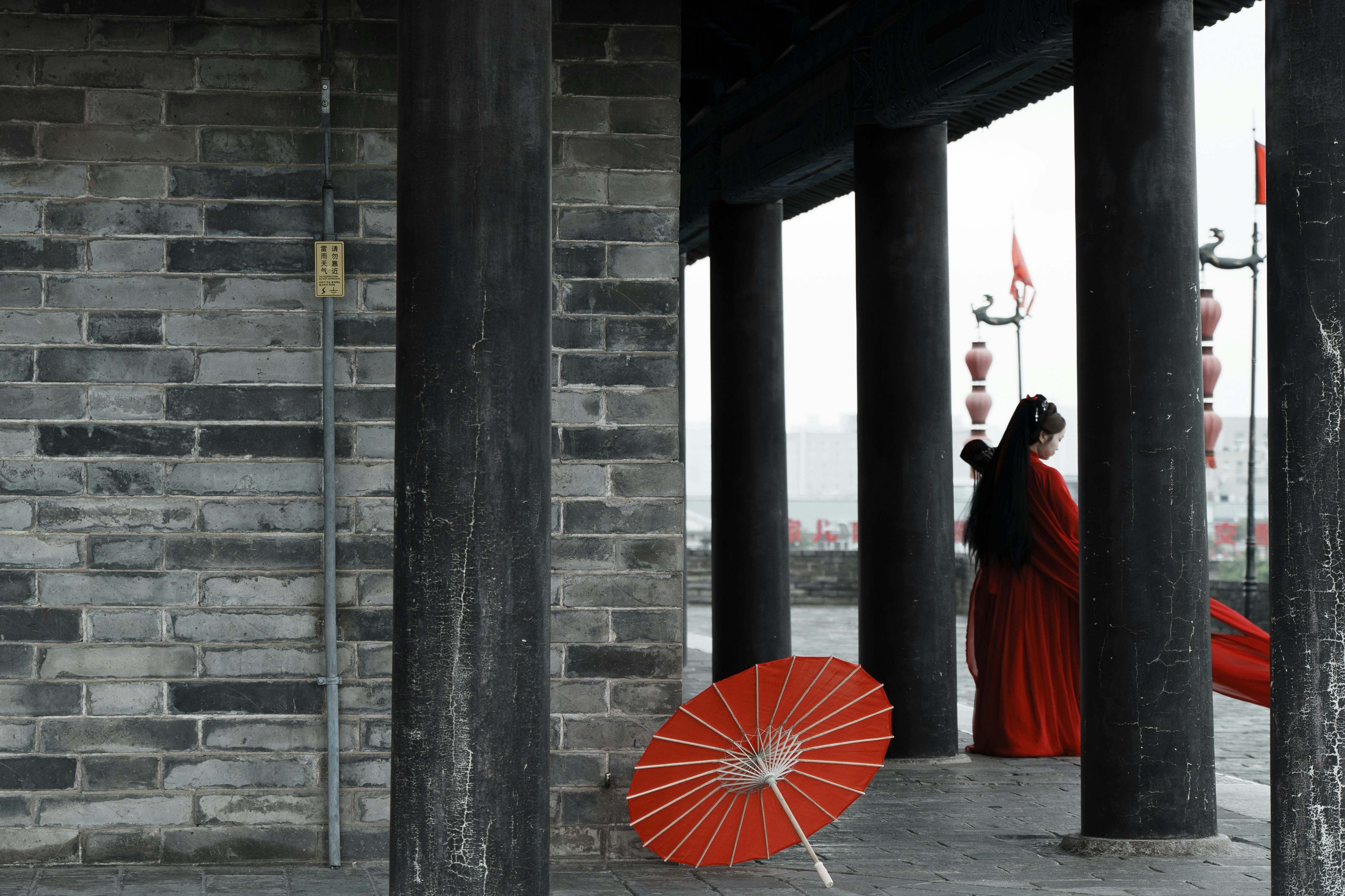 A woman in a flowing red garment stands near stone pillars, holding a red parasol, evoking a sense of cultural heritage and elegance.