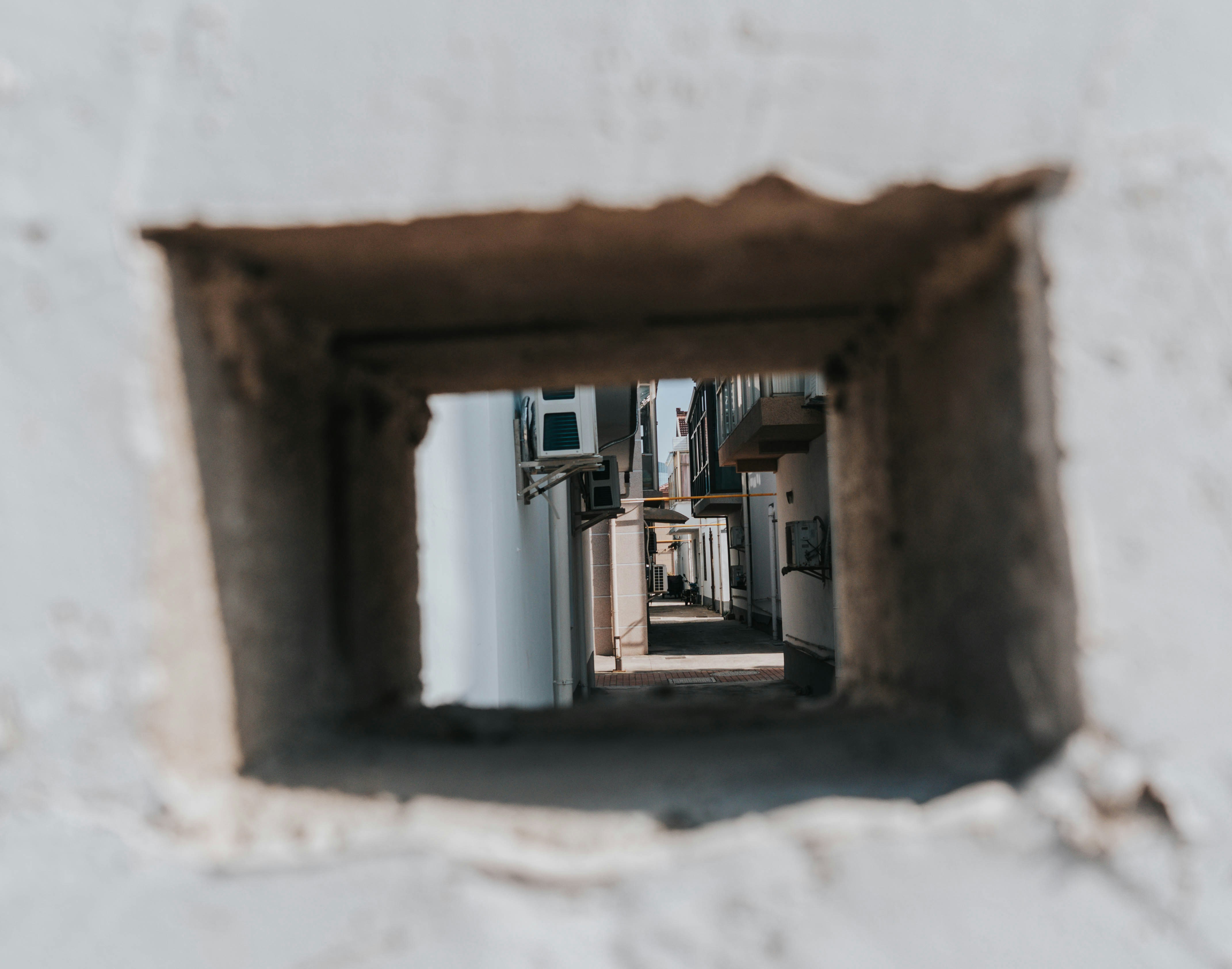 View through a narrow opening revealing a corridor of buildings bathed in sunlight. The composition highlights the contrast between the rough texture of the wall and the smoothness of the scene beyond.