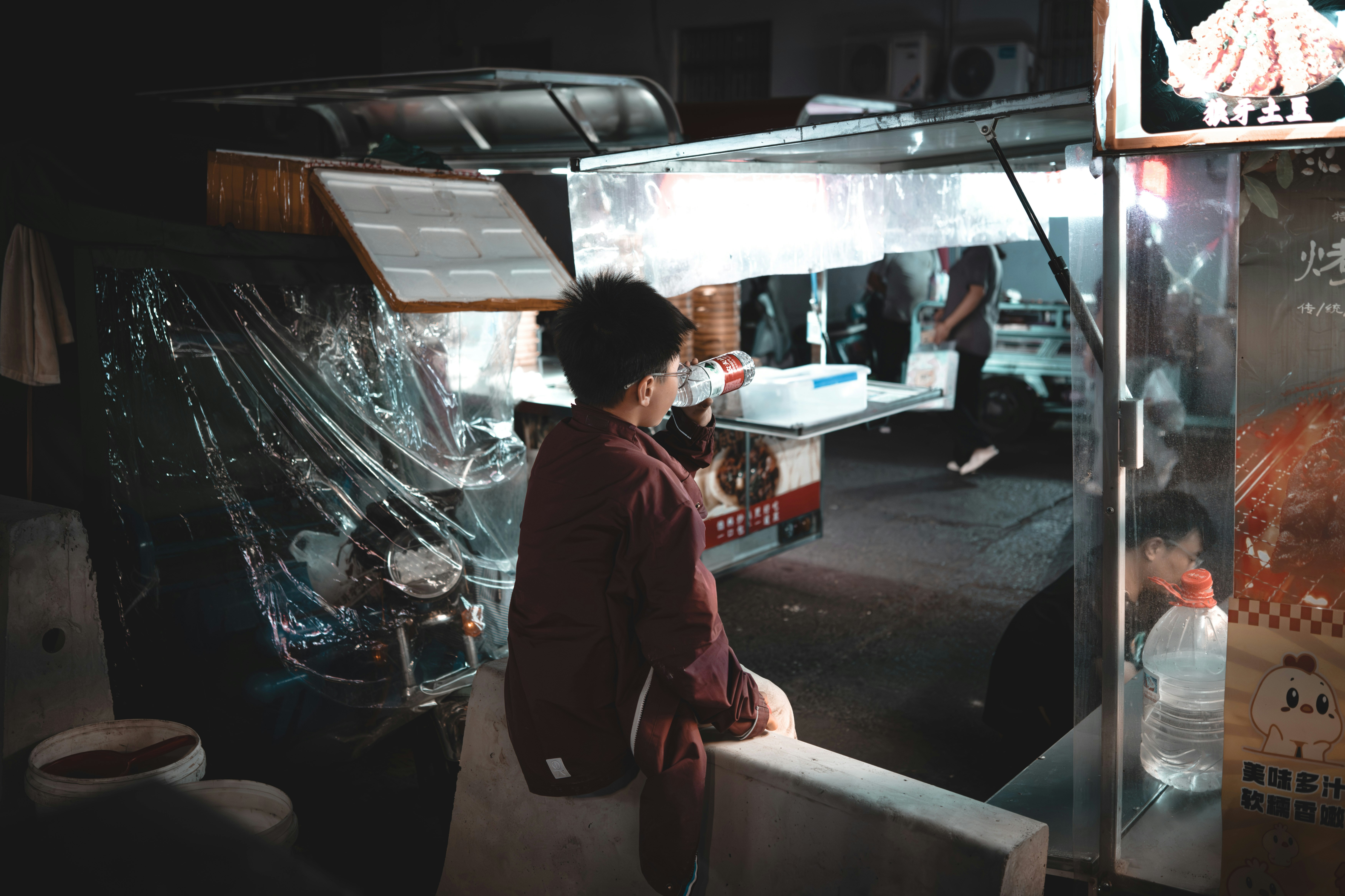 A young person sits contemplatively at a bustling night market, surrounded by food stalls and glowing lights. The scene captures the essence of urban life after dark.