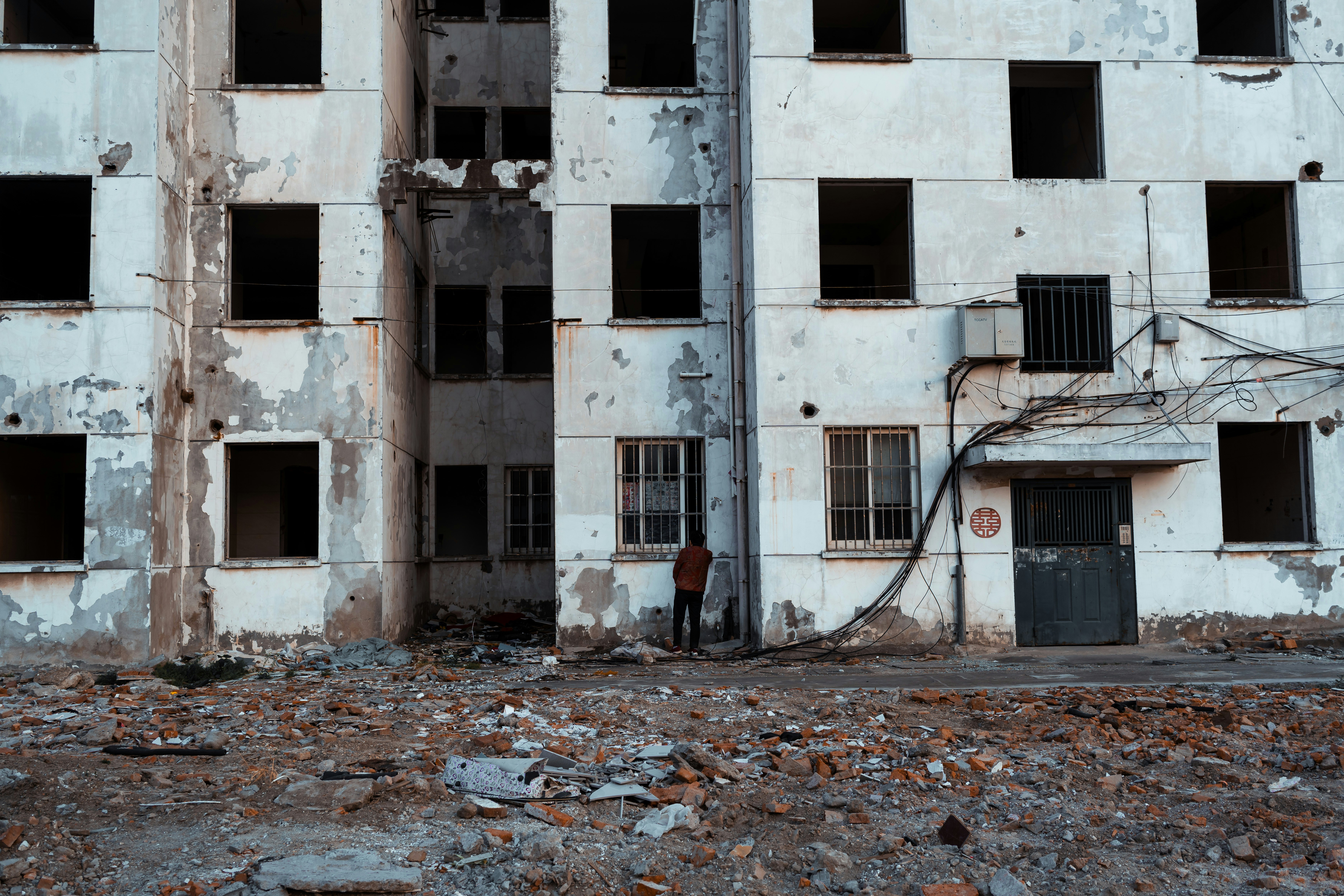 A lone figure stands before a damaged apartment building.