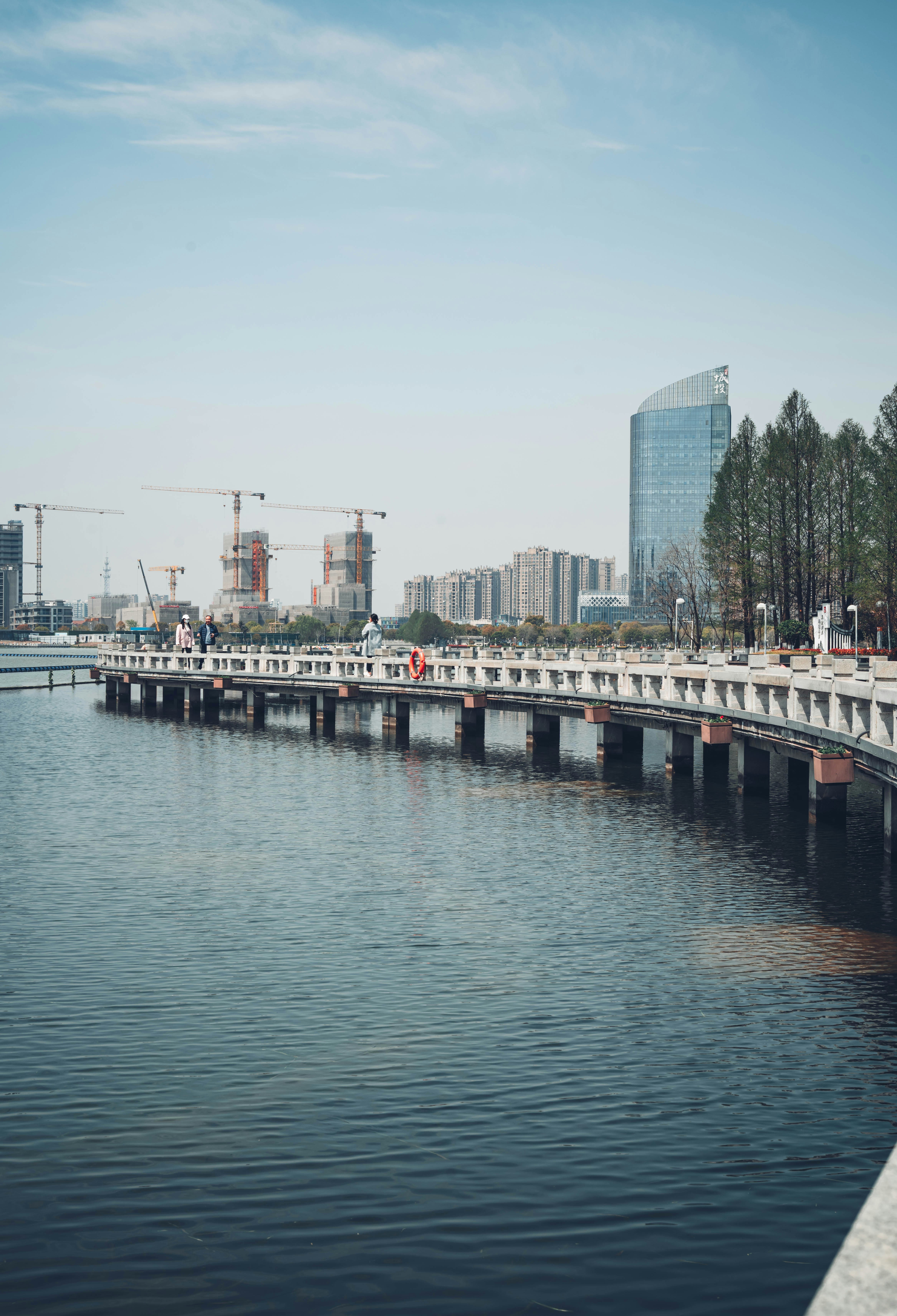 Curved walkway along a tranquil body of water, flanked by modern buildings and construction cranes in the background.