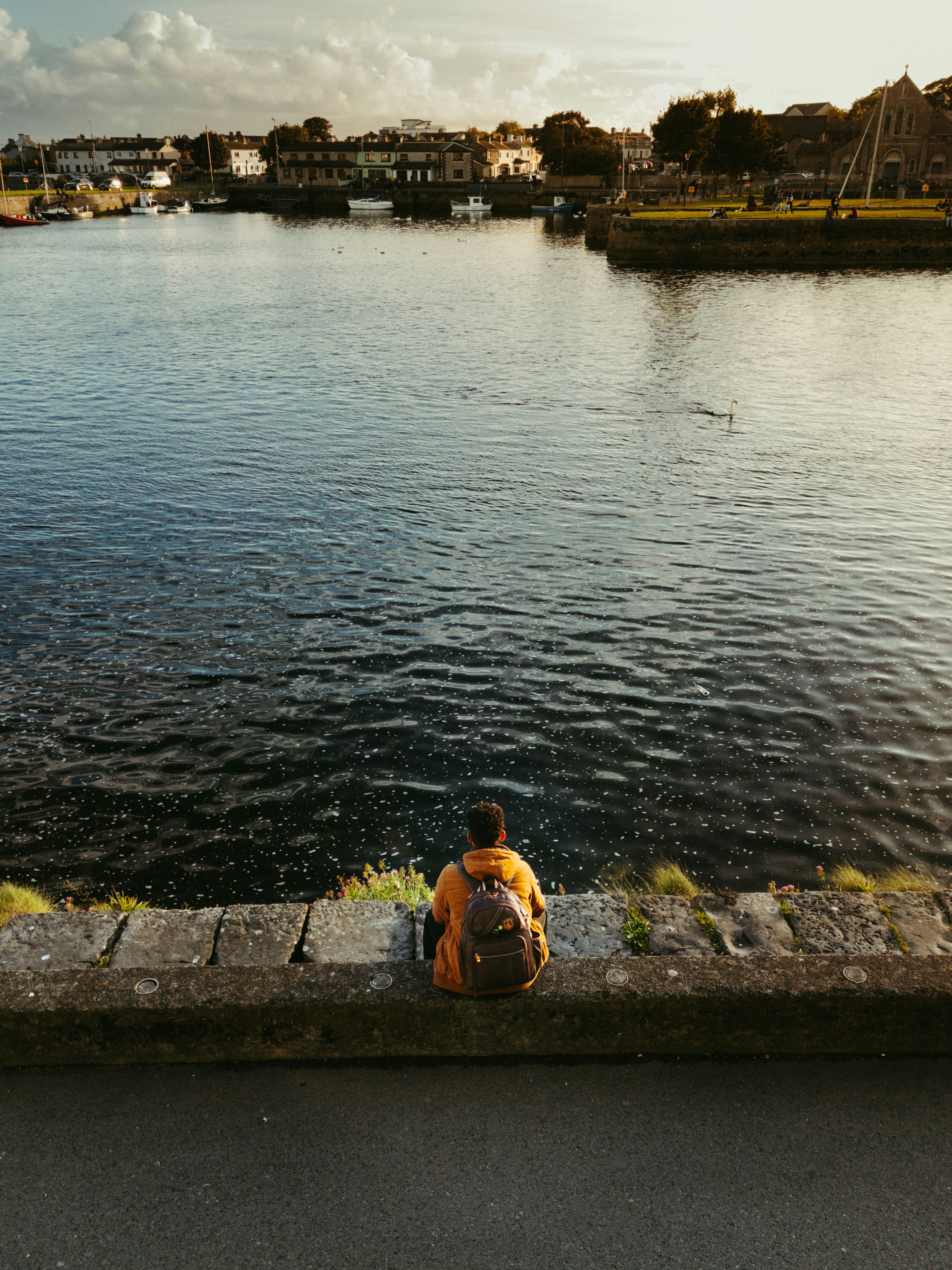 A person seated on a stone wall, gazing at the tranquil water, surrounded by a picturesque townscape. The scene captures a moment of reflection and connection with nature.