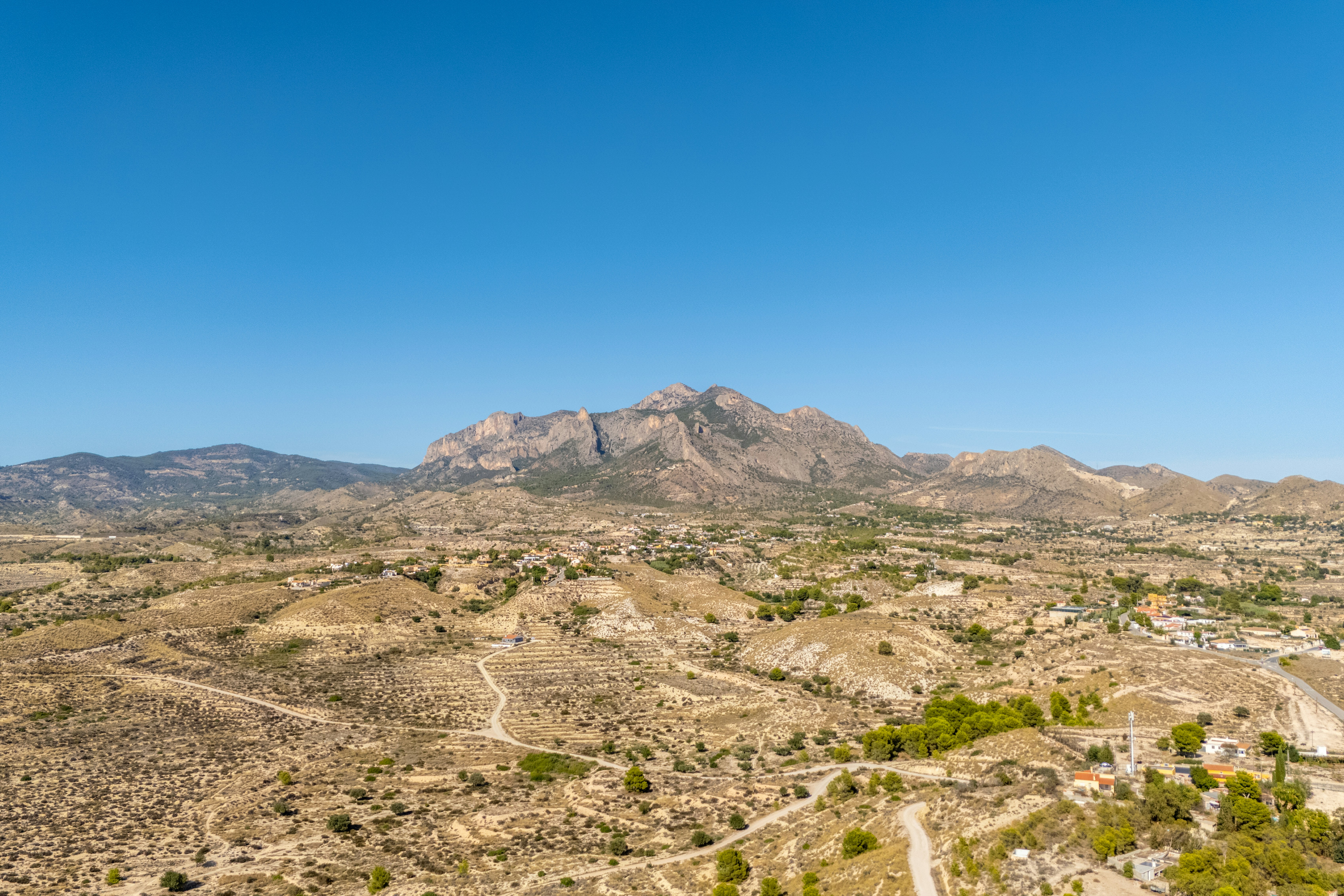 Expansive arid landscape with rolling hills and a prominent mountain range under a clear blue sky.