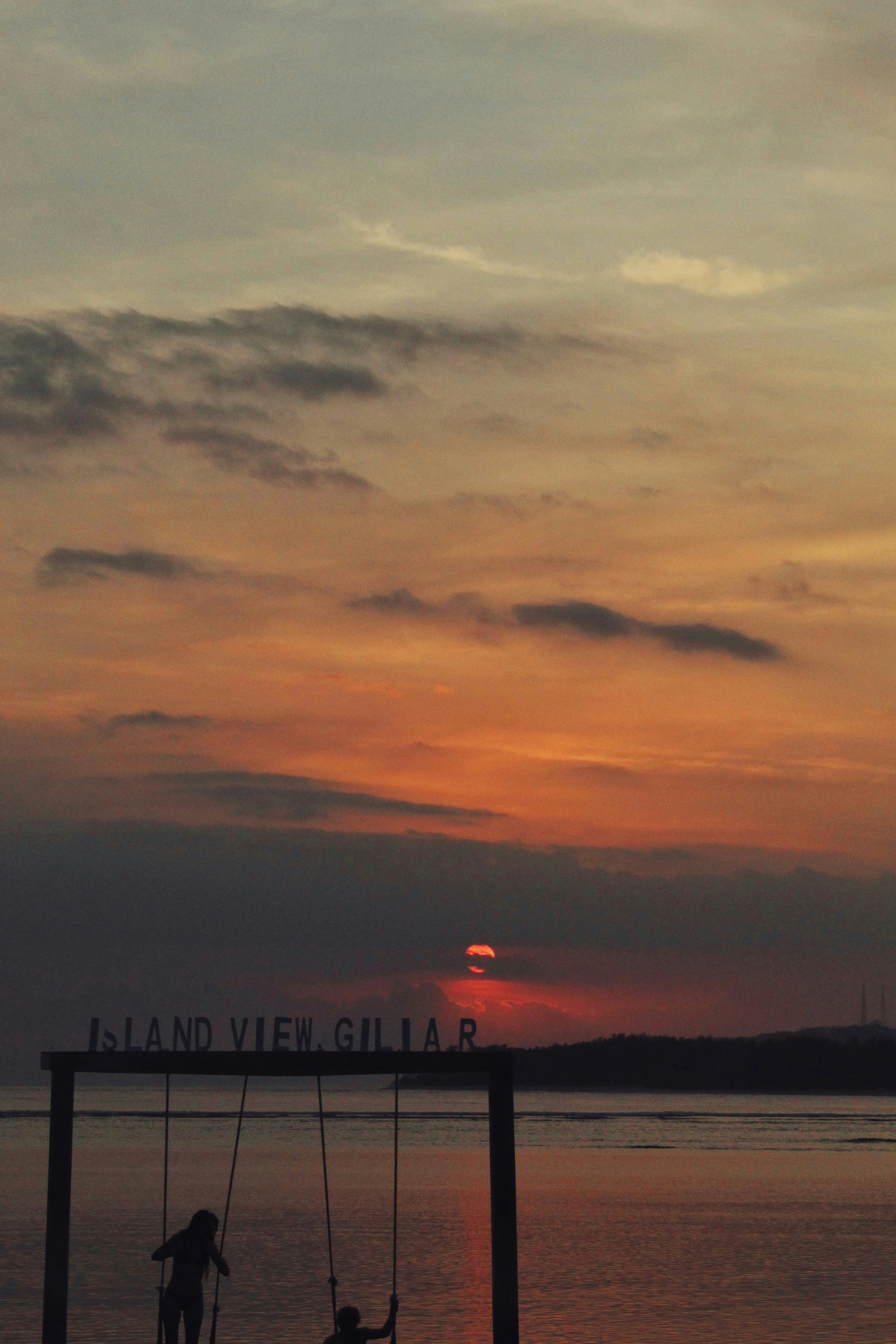 Two silhouetted figures on swings against a vibrant sunset, with the sign 'ISLAND VIEW GILI AR' prominently displayed above.