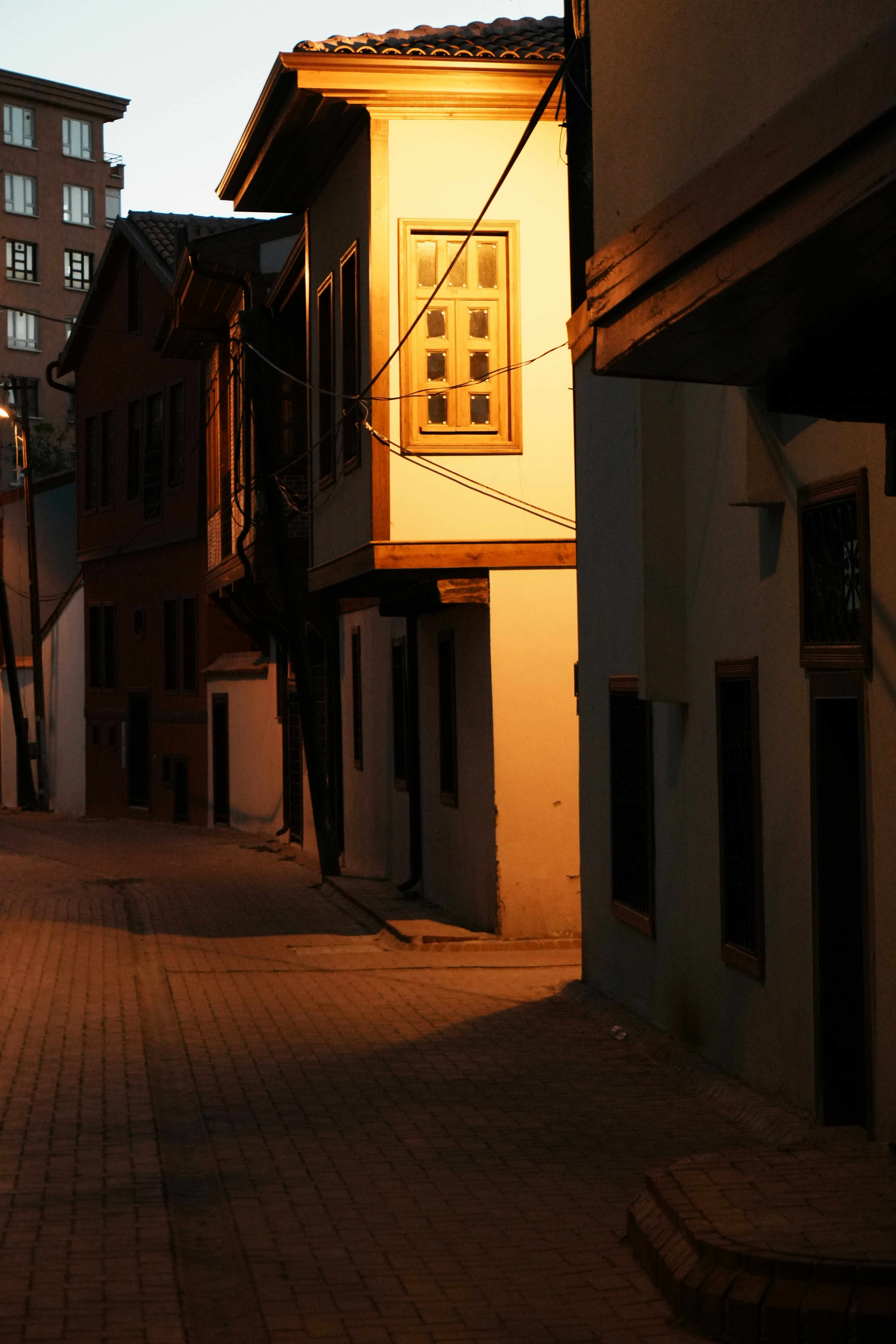 Title: Golden Hour on a Traditional Street Description: The last rays of a setting sun illuminate the facade of a traditional Turkish house, casting long shadows down a cobblestone street. The warm light highlights the historic architecture, creating a nostalgic atmosphere. | Narrow street lined with traditional houses at dusk.