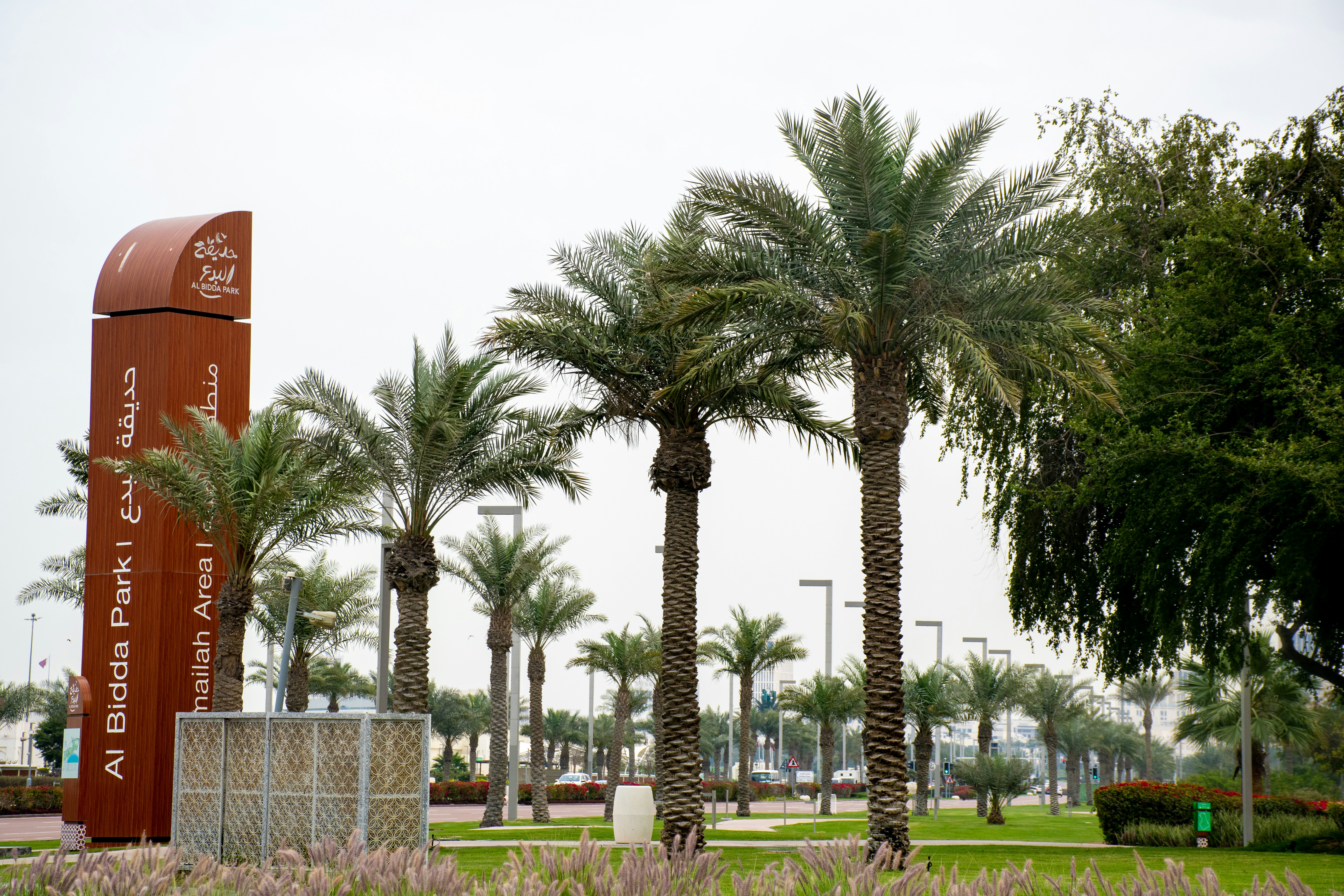 Signage for Al Bidda Park surrounded by palm trees and lush greenery in a tranquil urban setting.