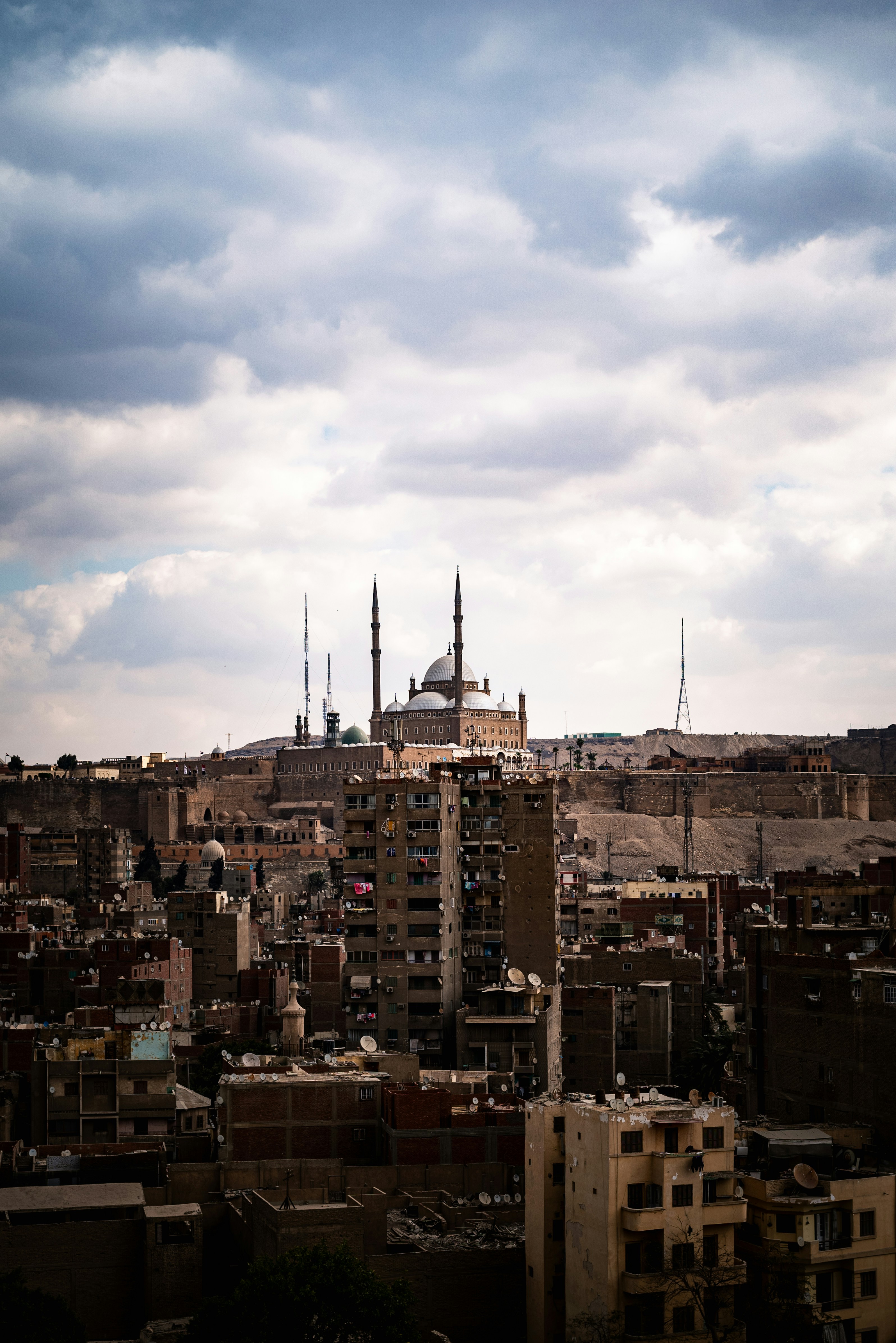 The majestic Citadel of Cairo looms over a bustling urban landscape, framed by dramatic clouds. The scene captures the juxtaposition of ancient architecture against modern city life.