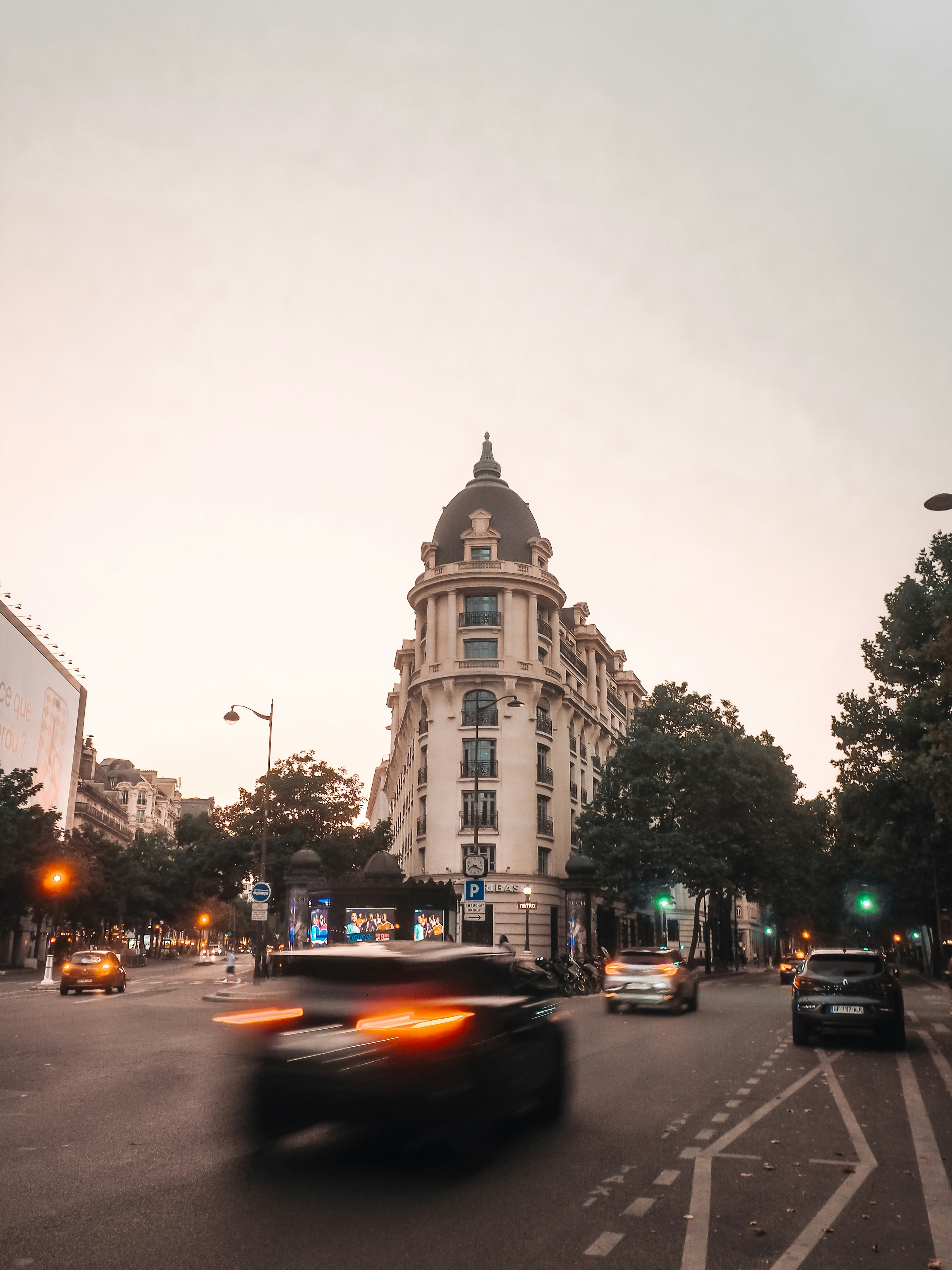 The Paris night street scene comes alive with the Eiffel Tower glowing above. This image merges street photography, travel romance, honeymoon vibes, and Parisian lifestyle. A couple sits together in a candid street moment, surrounded by urban night lights, city reflections, and golden atmosphere. It’s a story of romance, love, passion, tenderness, and intimacy within the urban cityscape. Perfect for wedding inspiration, anniversary trips, engagement photos, and romantic travel campaigns, this photo proves why Paris is the eternal city of love and why street photography finds magic in every cor | Cars driving past a building at dusk.