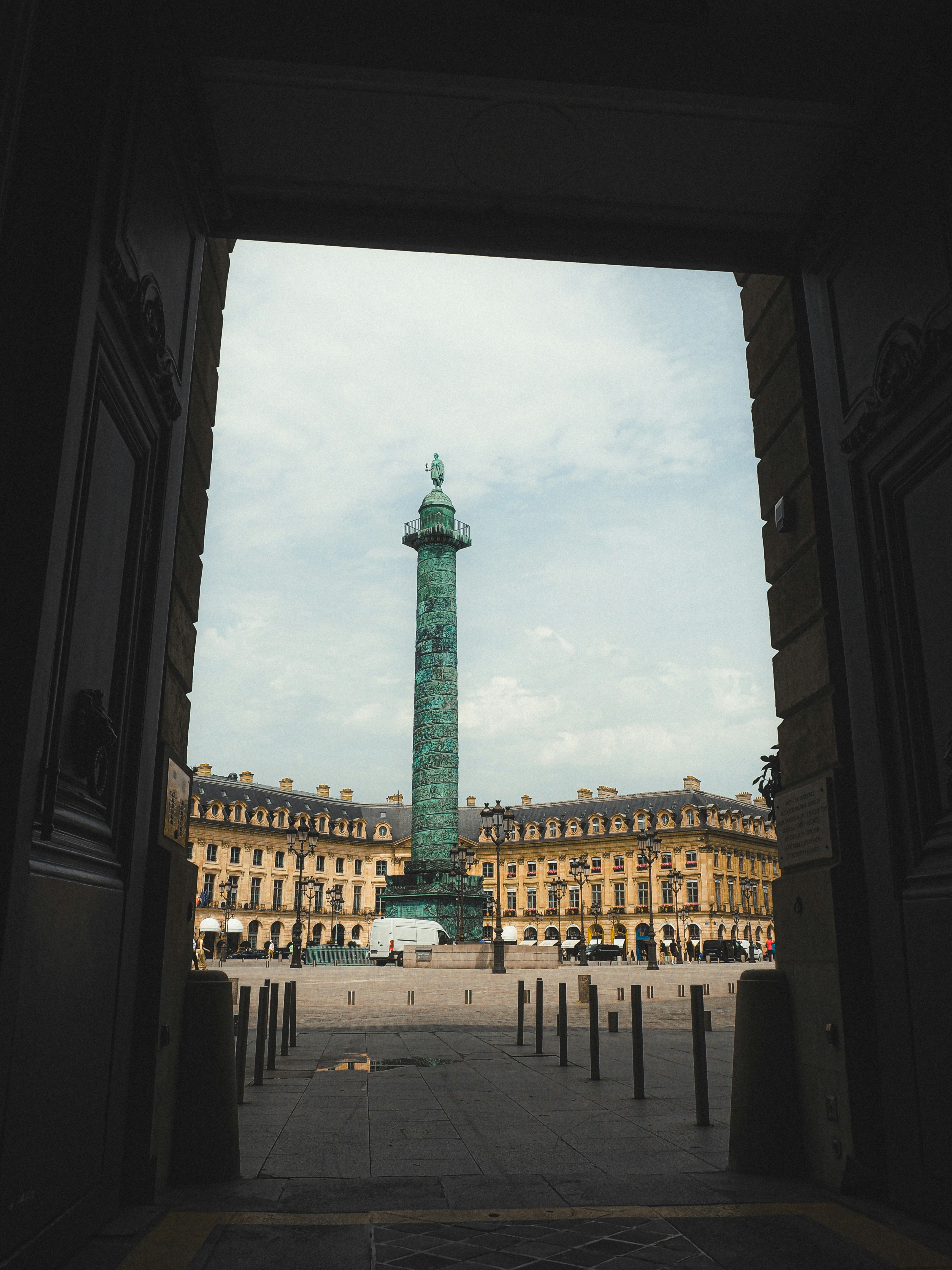 View through an ornate doorway framing the Column of Vendôme, surrounded by historic architecture and a cloudy sky.