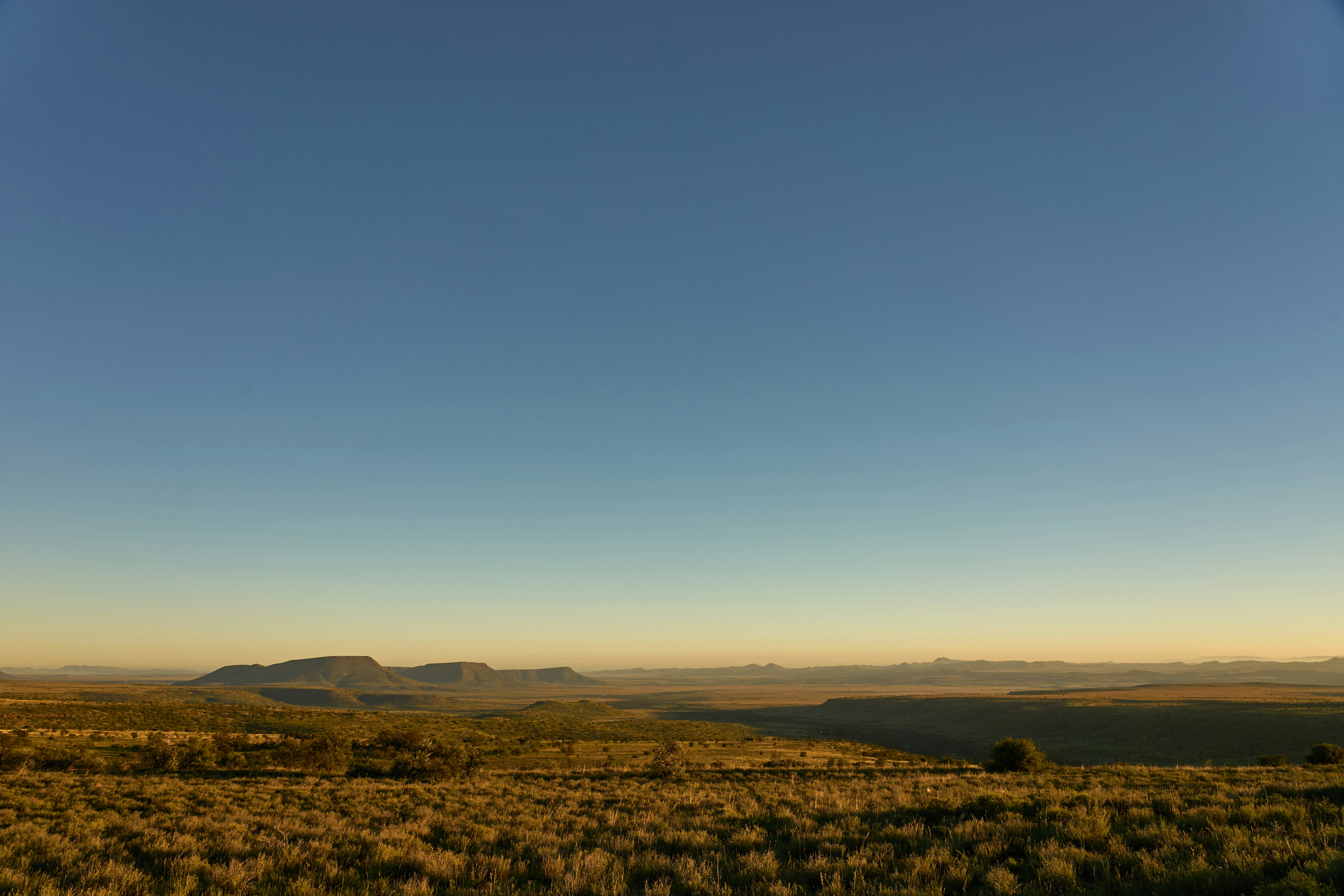 Vast landscape under a clear sky, featuring rolling hills and distant mountains bathed in soft light. The scene captures the tranquil essence of nature's expanse.