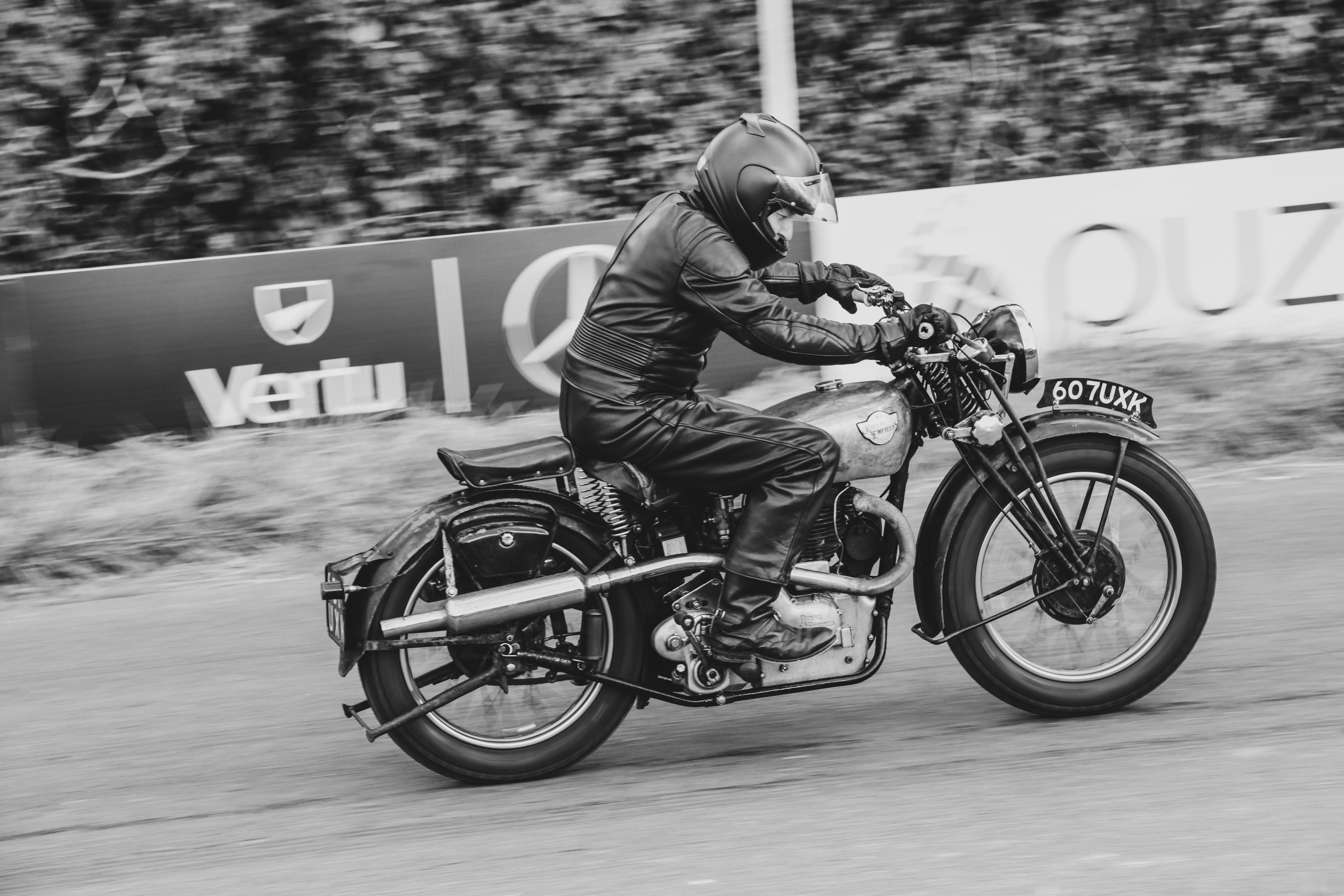A motorcyclist in leather gear speeds down a track on a classic motorcycle, captured in dynamic motion. The scene is presented in black and white, emphasizing the nostalgia of the moment.