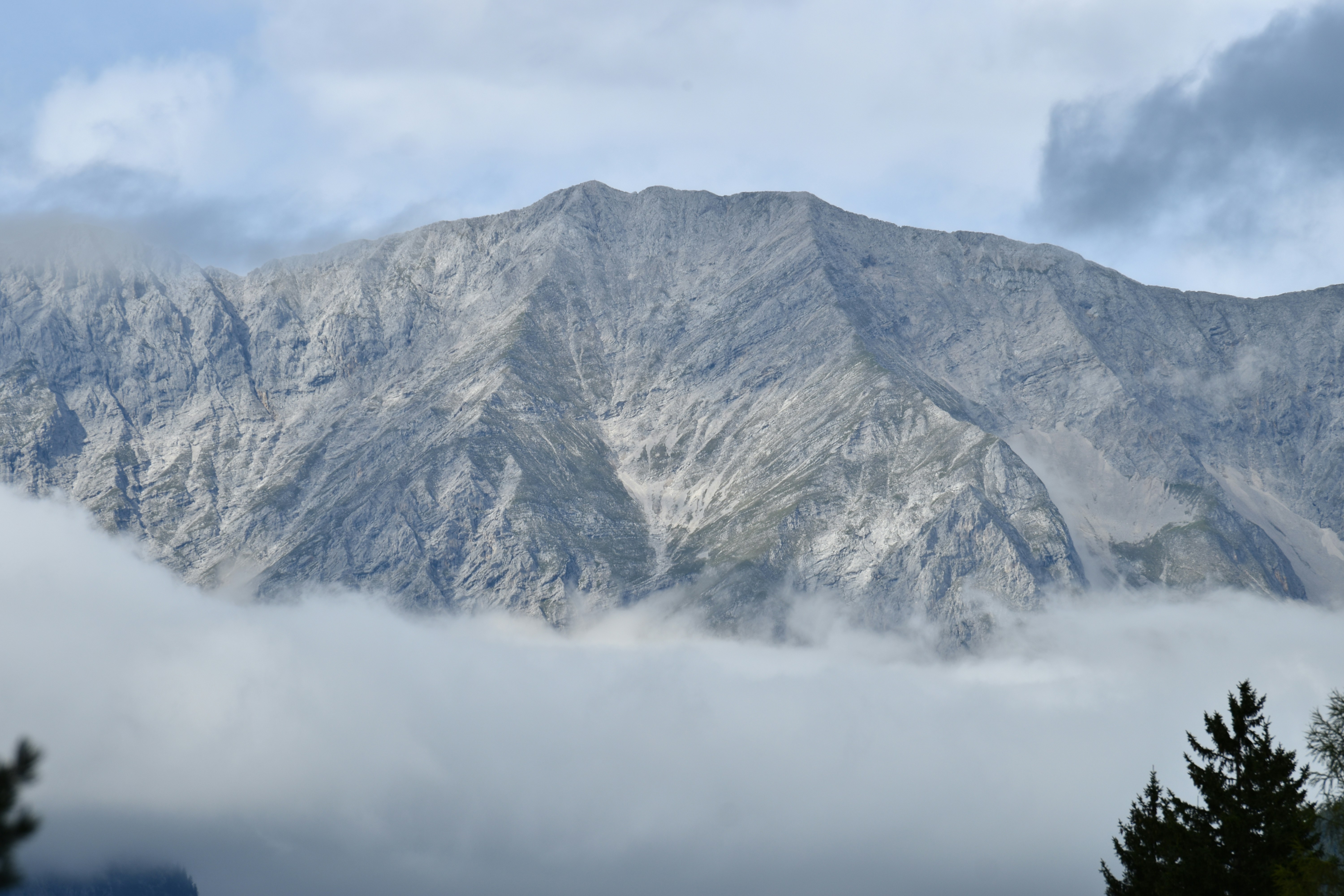 Majestic mountain peaks rise beneath a blue sky with light clouds. A serene and imposing landscape, ideal for projects related to nature, adventure, and tranquility. | Majestic mountain peak emerges from swirling clouds