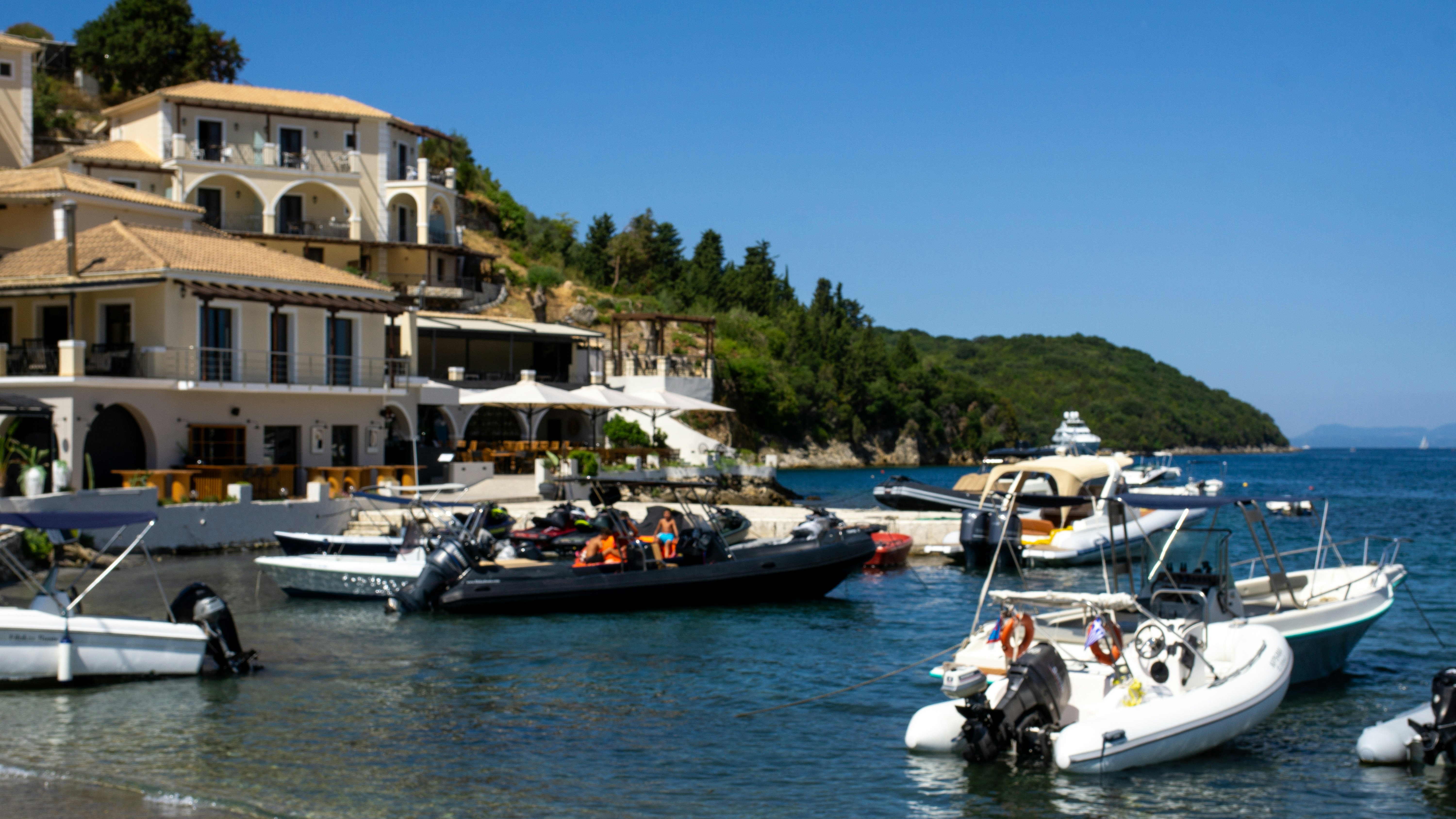 Boats anchored in a tranquil harbor beside a charming waterfront restaurant, framed by lush greenery and clear blue skies.