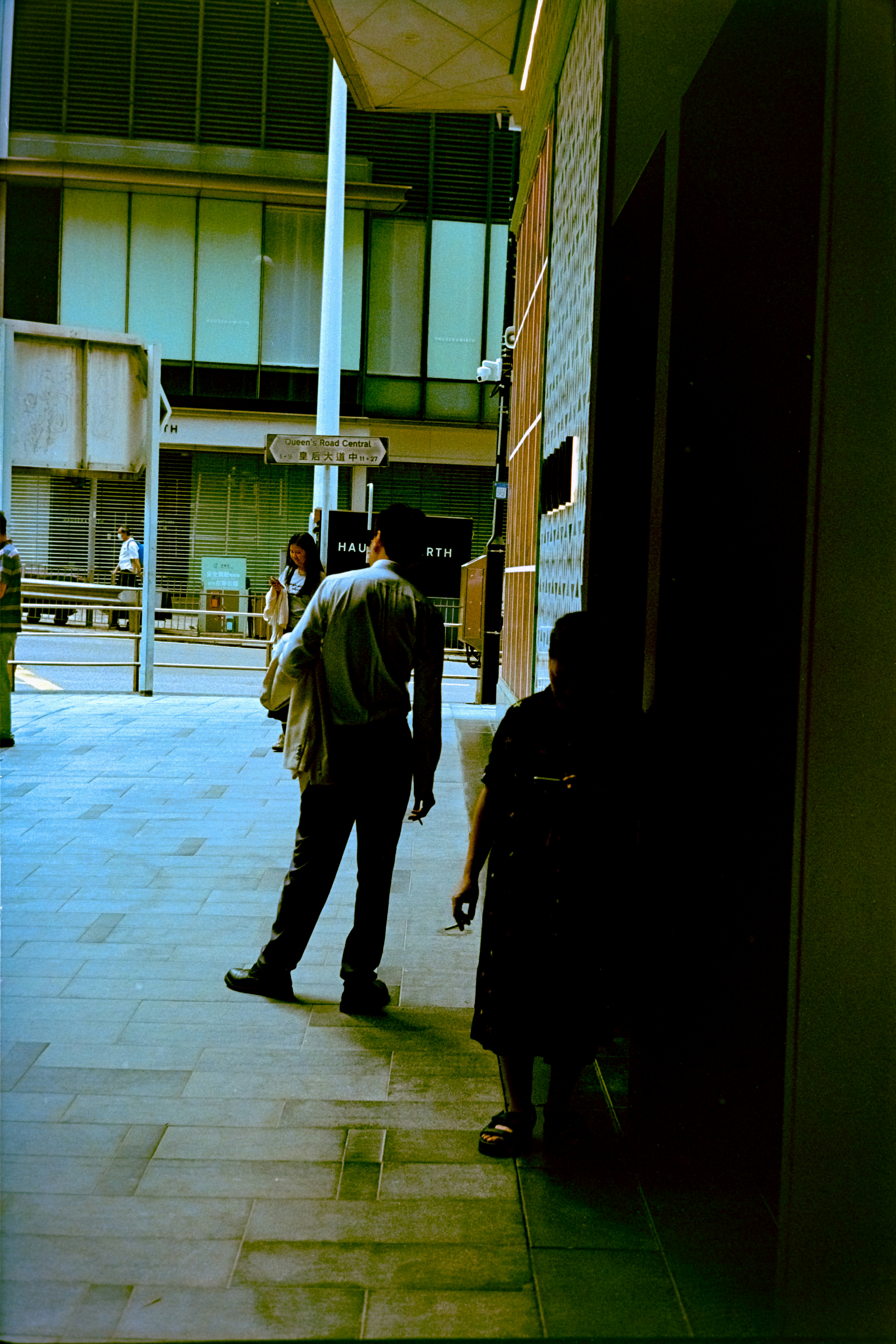 A man stands in a dimly lit corridor, casting a shadow, while a woman approaches from the side. The interplay of light and shadow creates a sense of depth.