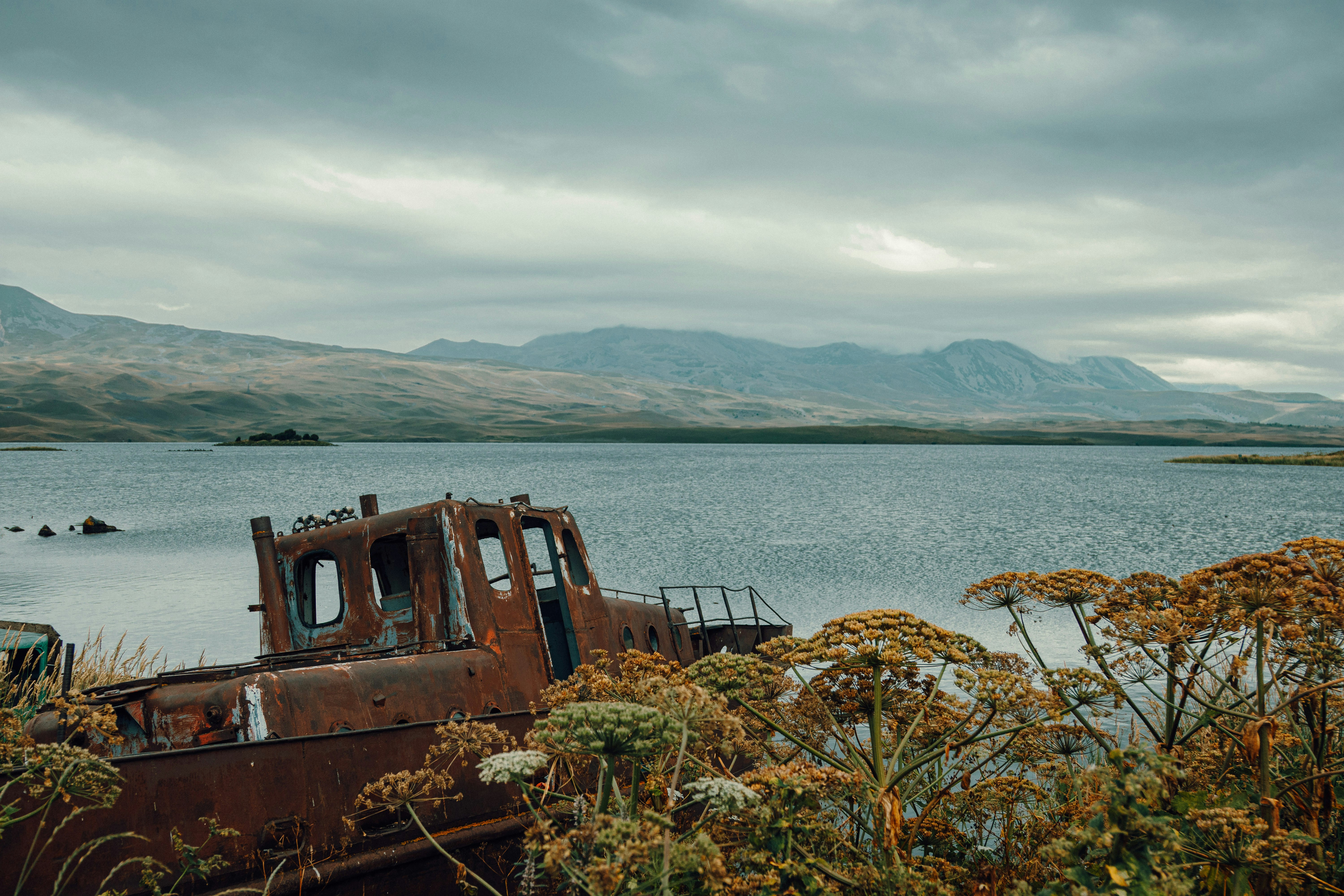 An abandoned old rusty boat on the shore of a mountain lake under a cloudy autumn sky in a remote corner of a national park in Georgia near Borjomi. | Rusty shipwreck on a serene lake with mountains.