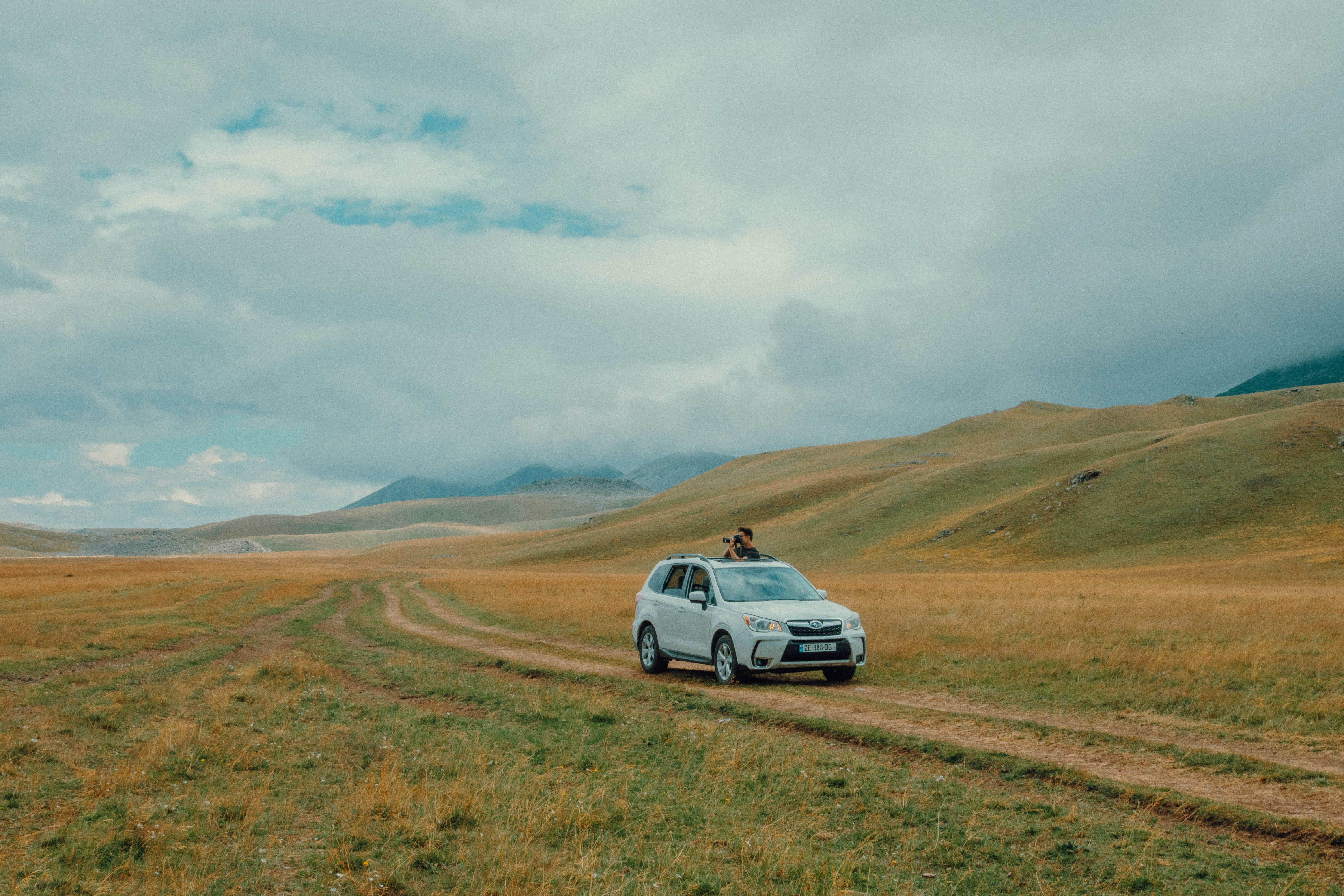 A white SUV navigates winding dirt tracks across expansive golden grasslands under a moody sky.