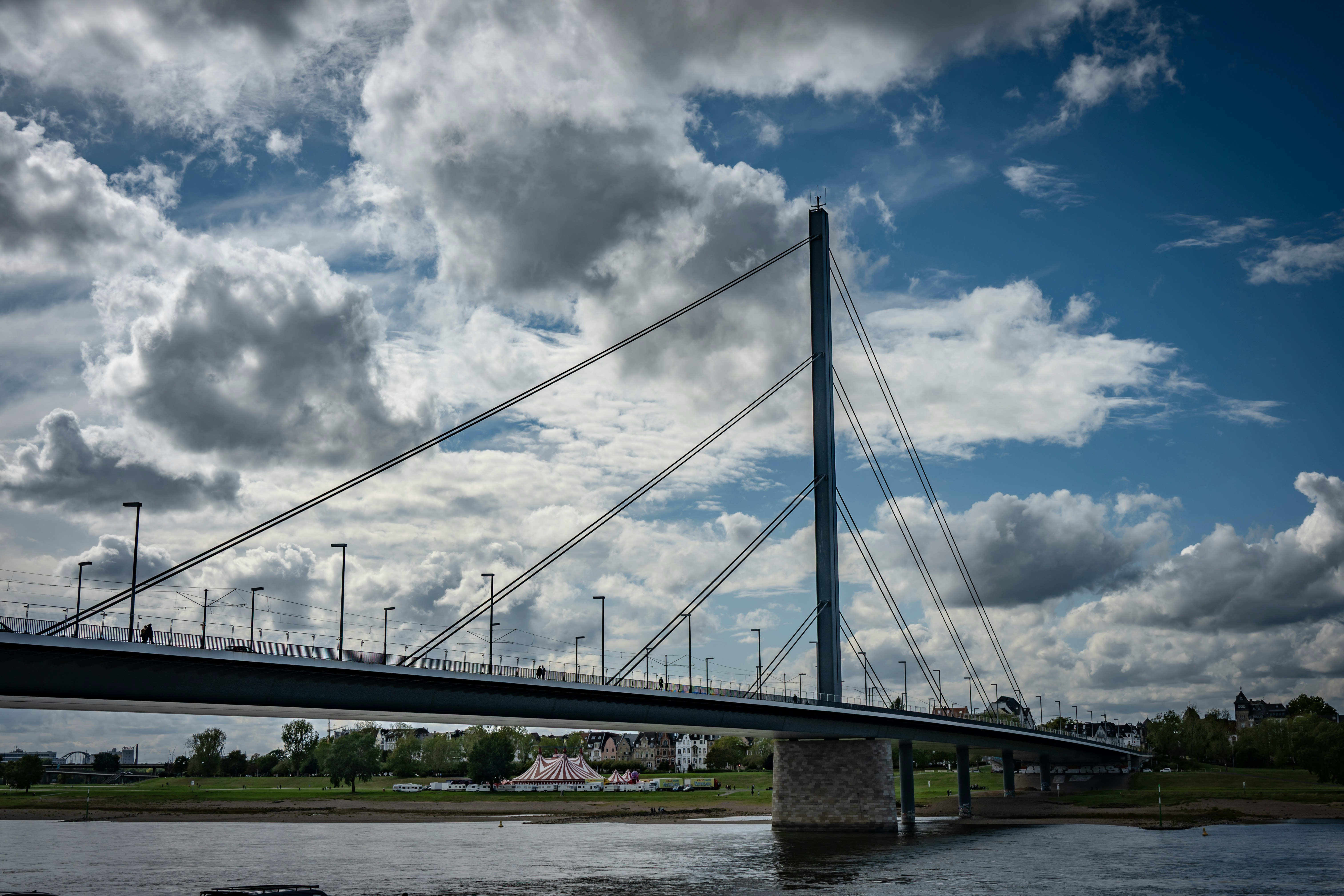 A sleek cable-stayed bridge spans across a river, framed by a dynamic sky filled with clouds. The scene captures the intersection of nature and modern architecture.