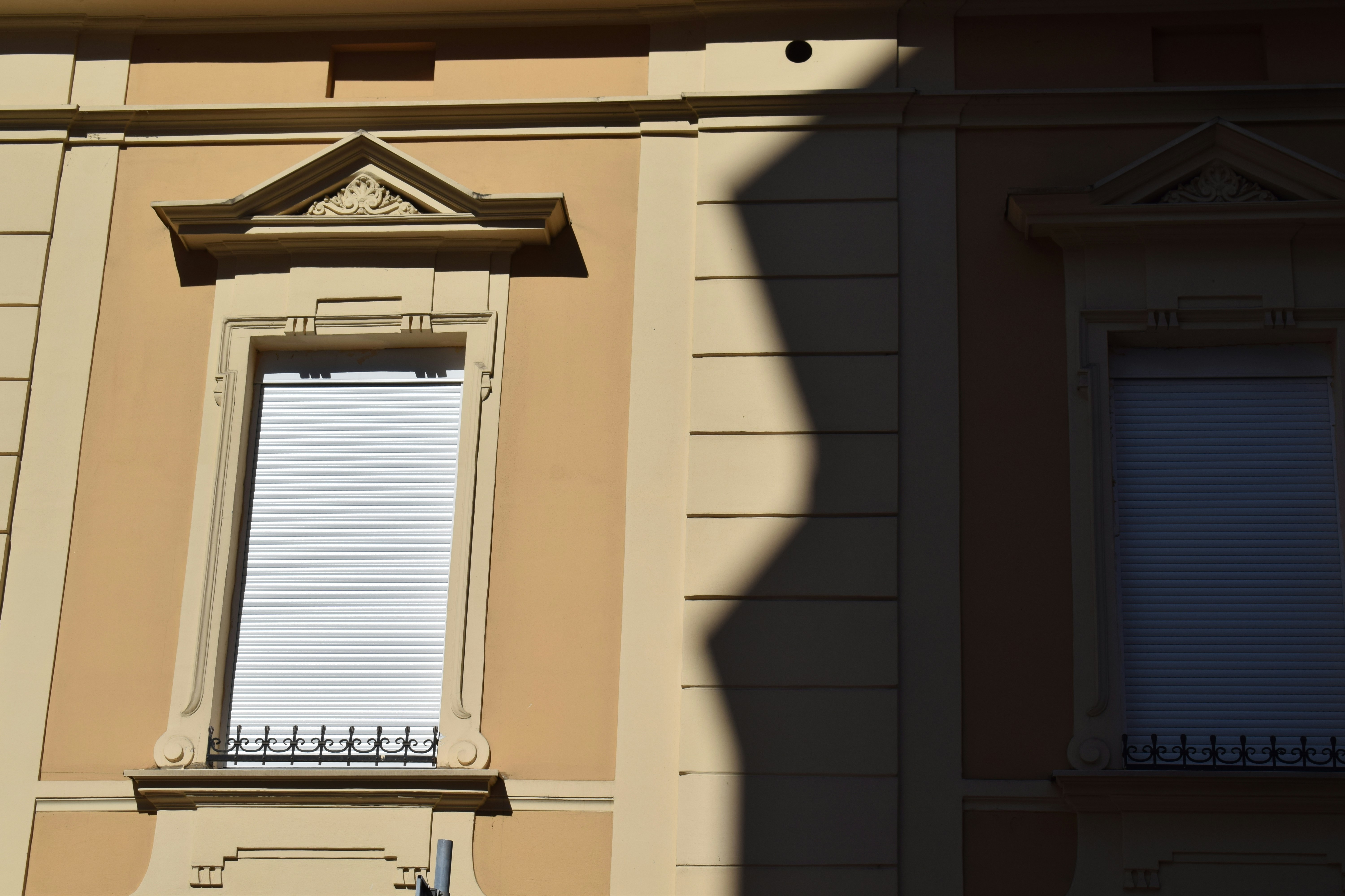 Elegant facade featuring two closed window shutters, with intricate architectural details and a pronounced shadow cast across the wall.